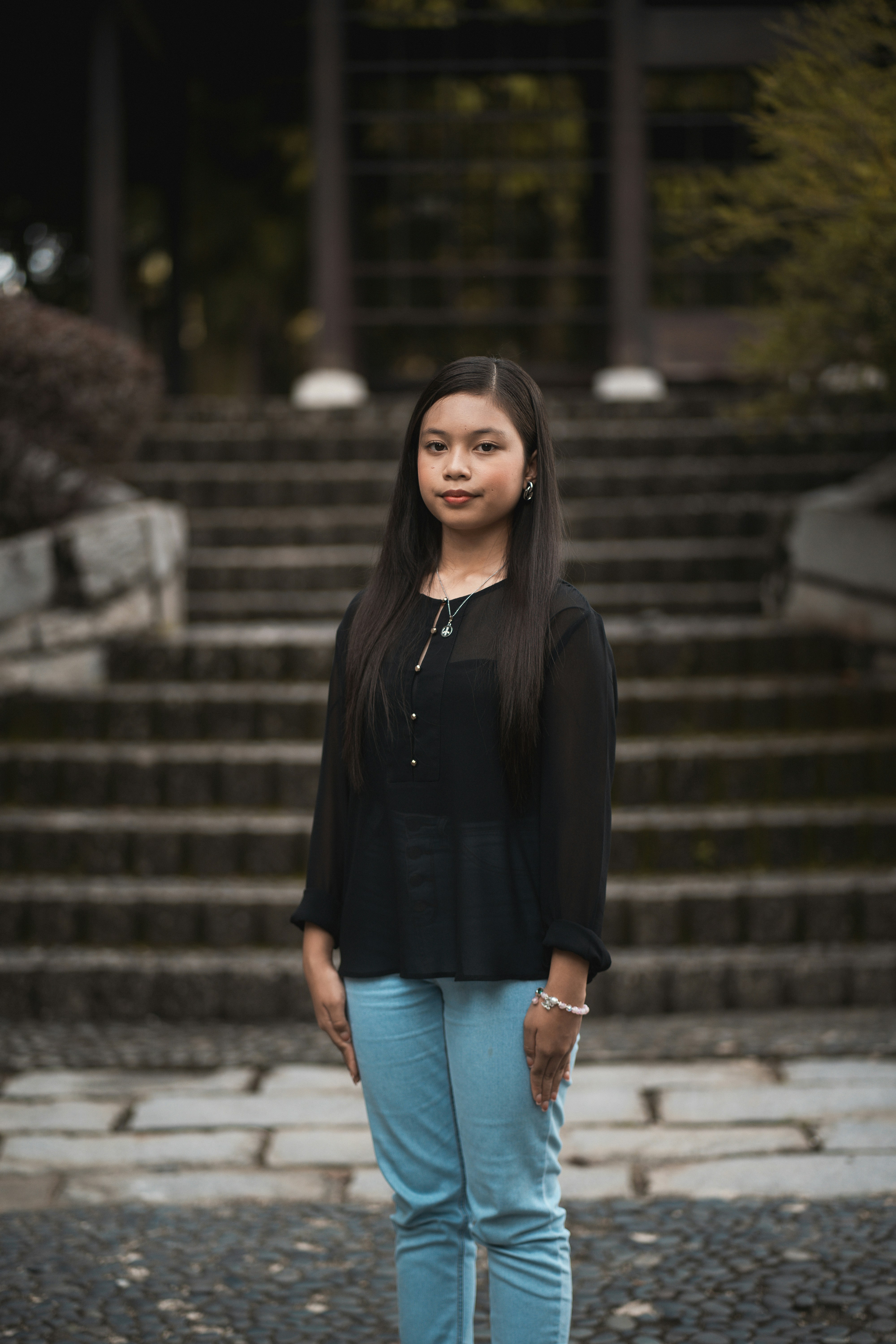 Young woman standing on a stone pathway with blurred steps and greenery in the background.