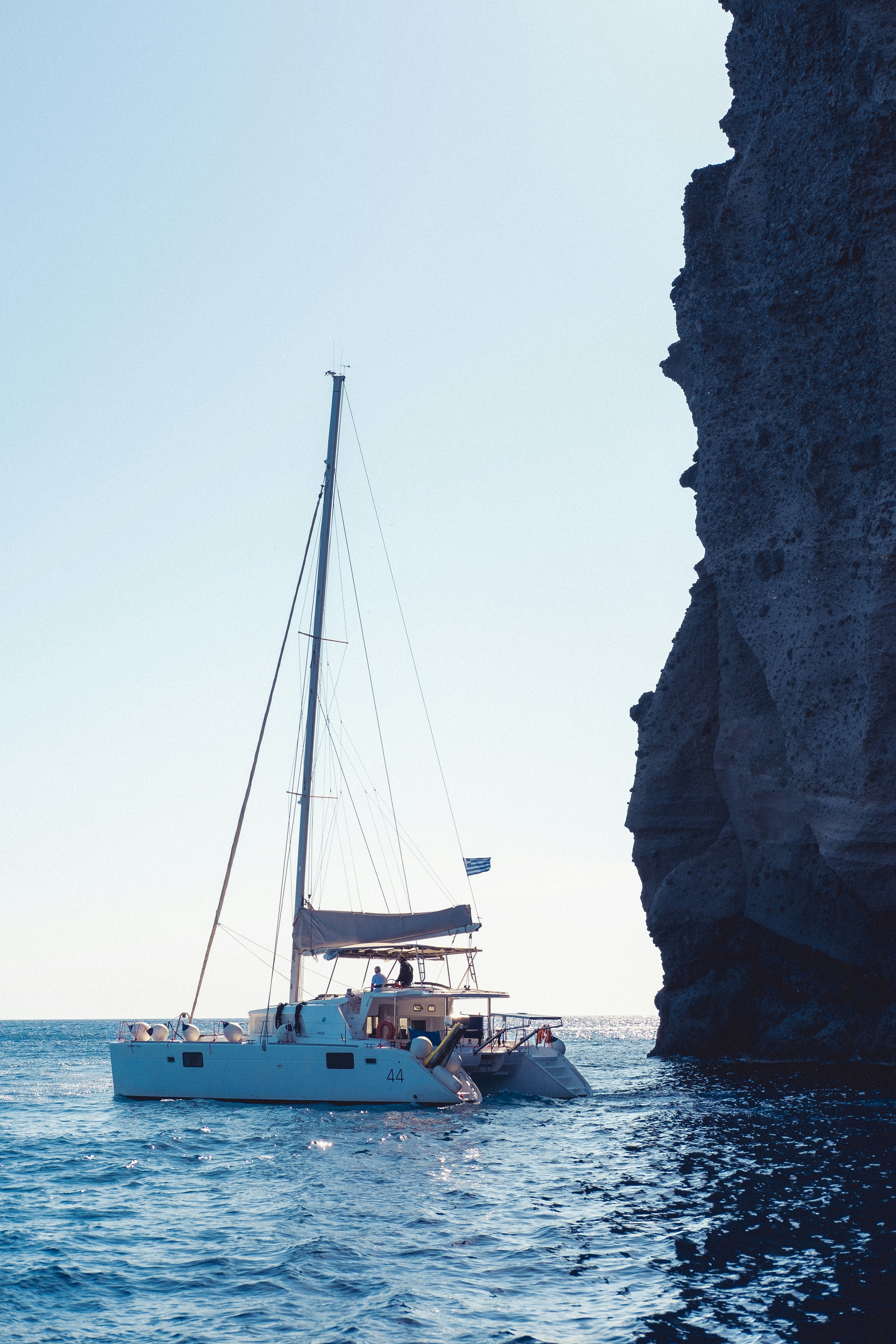 A serene sailing yacht anchored in calm blue waters near a towering cliffside. The yacht features white sails and a sleek design, perfectly capturing the essence of maritime adventure. The towering cliff adds dramatic contrast, highlighting the natural beauty and tranquility of the sea.