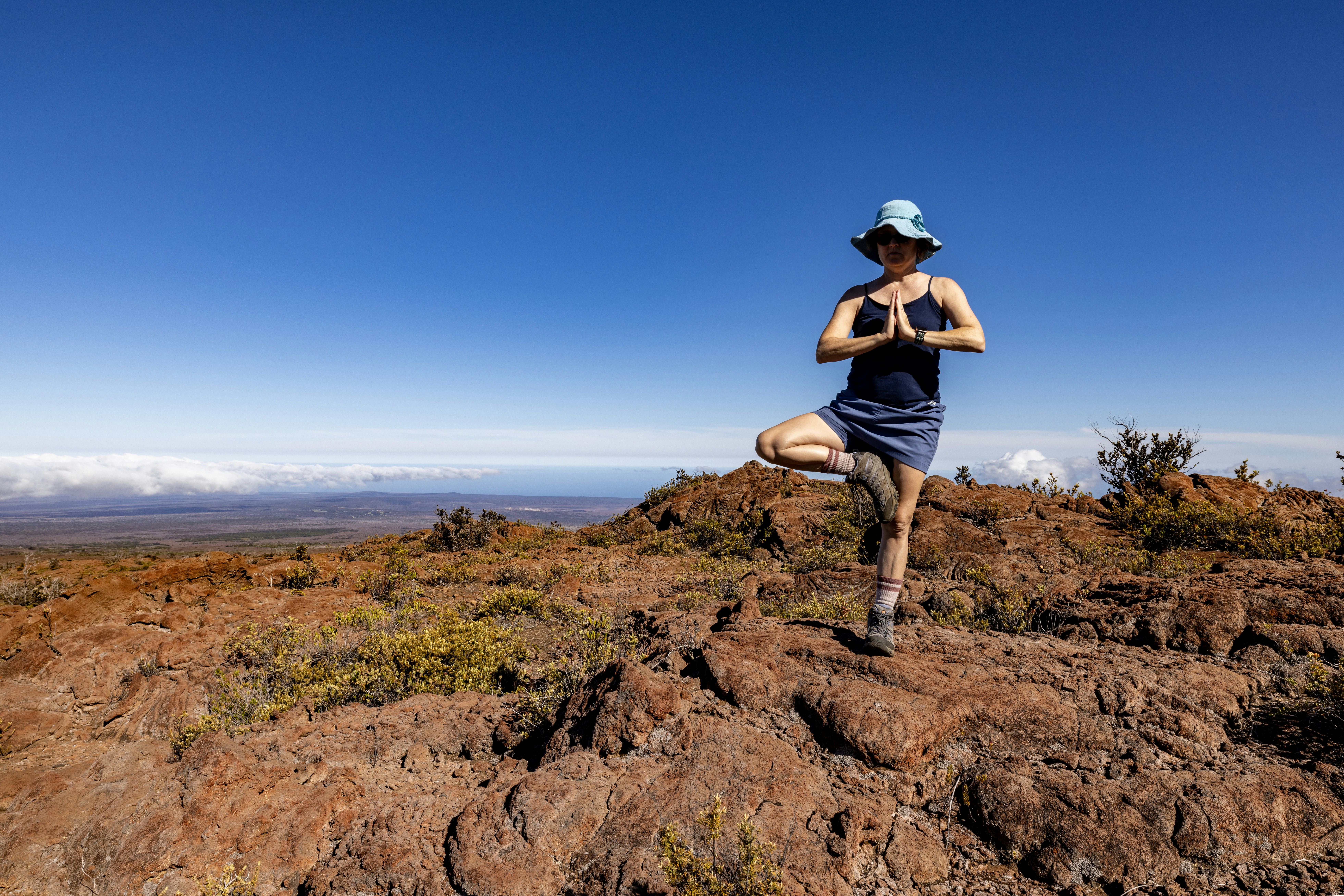 Person practicing yoga on rocky terrain with expansive sky and distant clouds on Mauna Loa Volcano.