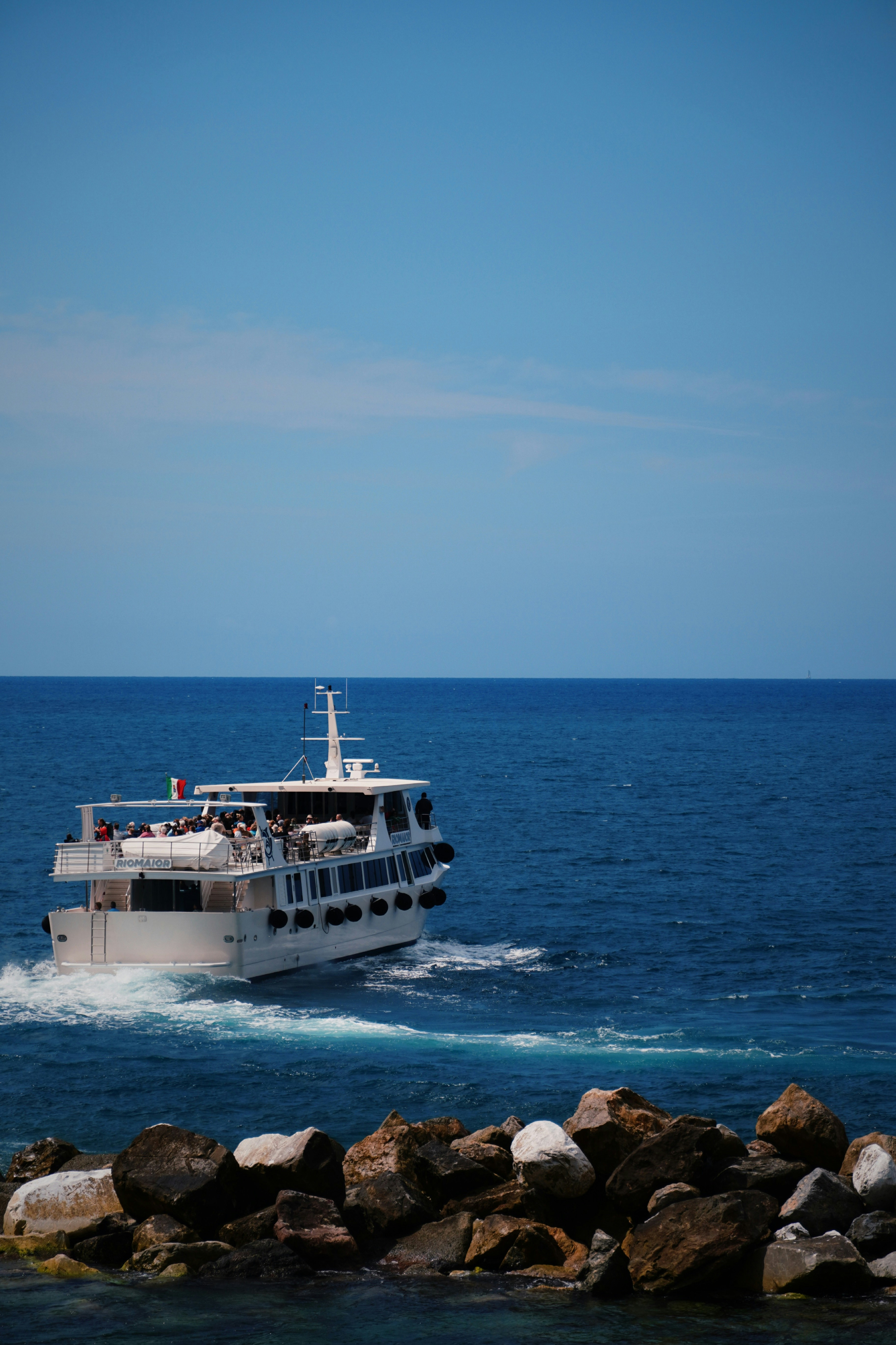A ferry boat with tourists against blue water, with waves and rocks in the foreground.