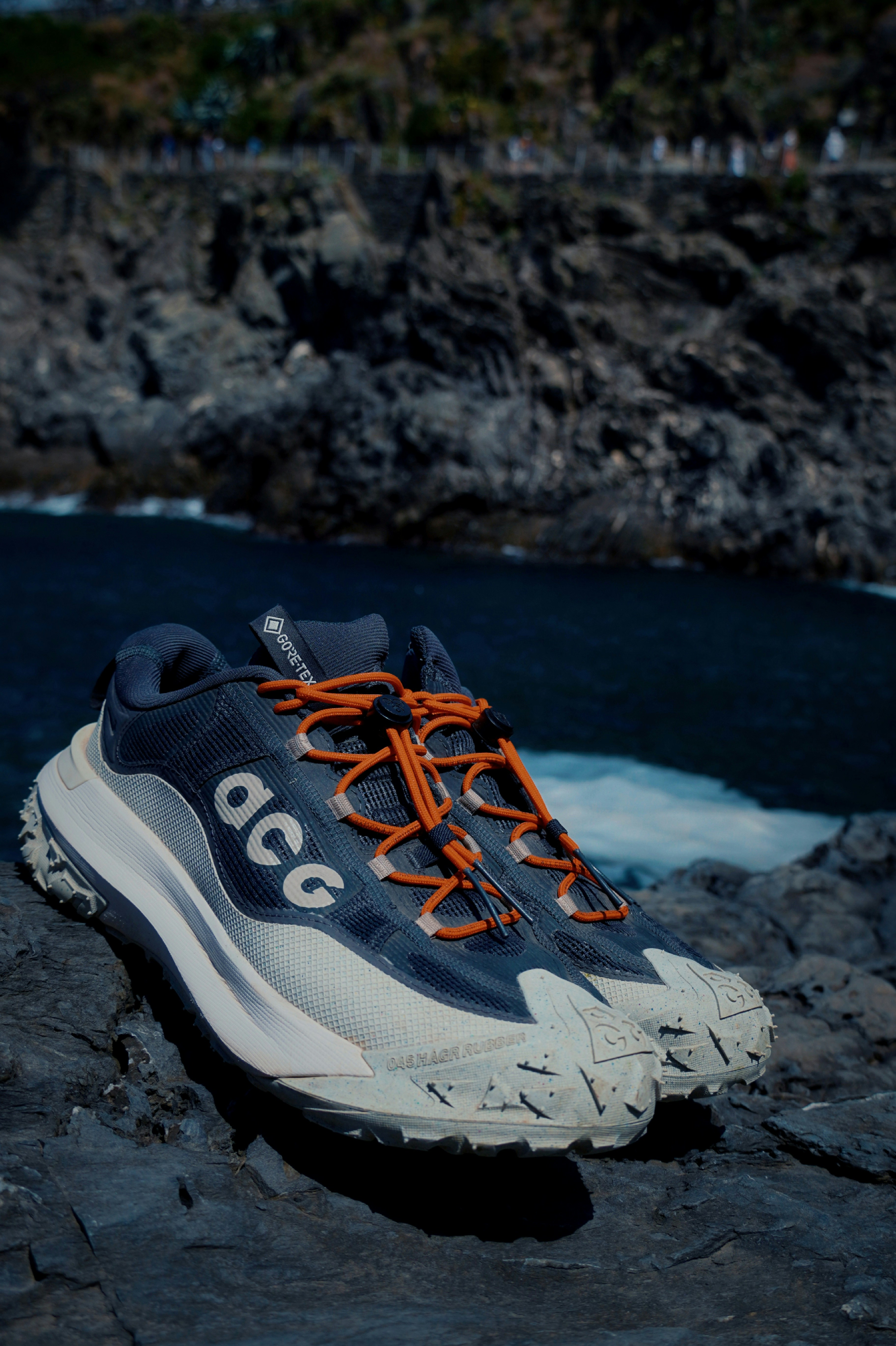 A pair of sneakers with orange laces against a rocky shore, with the sea and cliffs in the background.