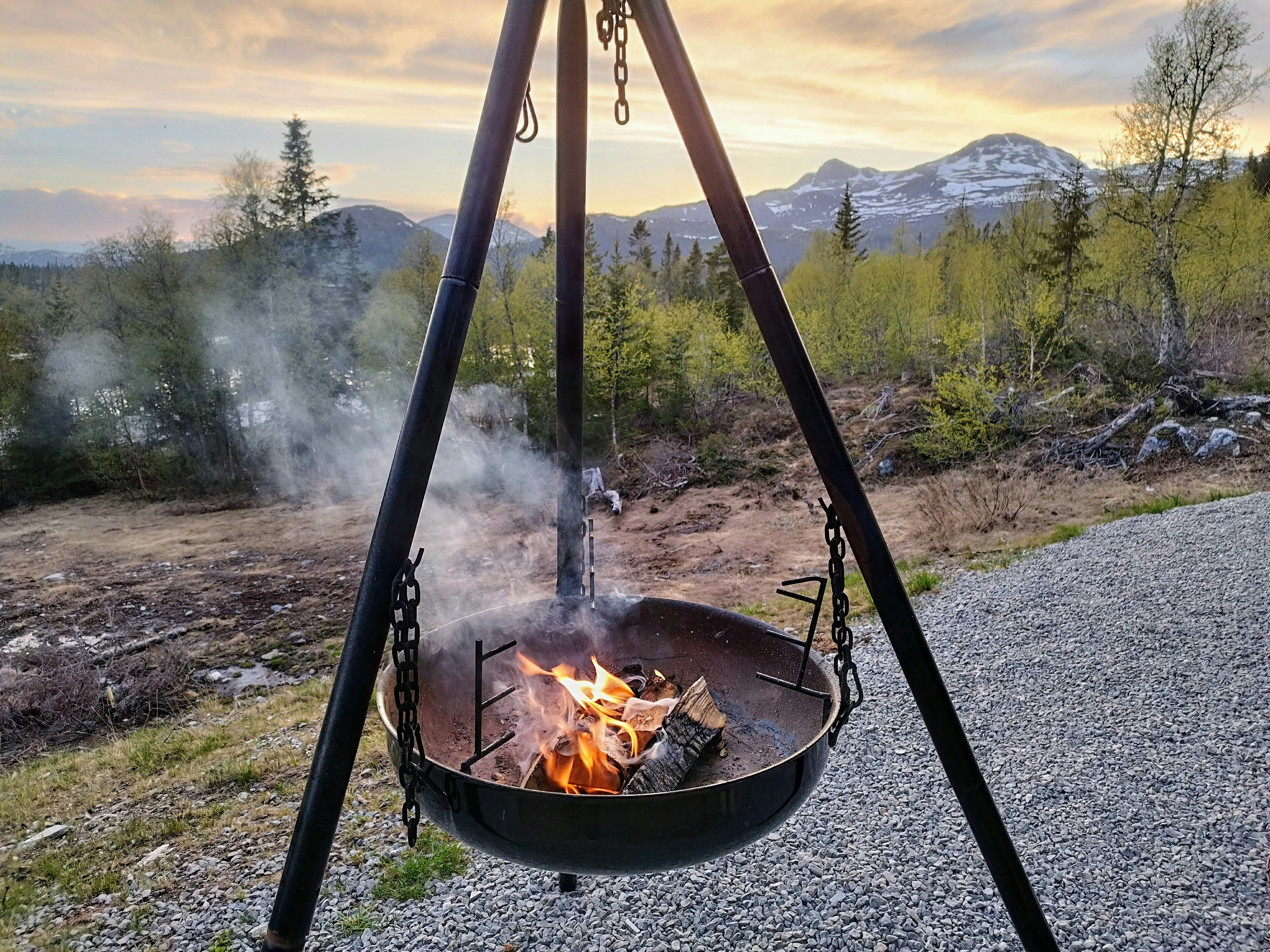 Tripod-hung cauldron over a crackling campfire at a mountain campsite, smoke curling upward. Alpine pines and snow-capped peaks glow in a warm sunset backdrop.