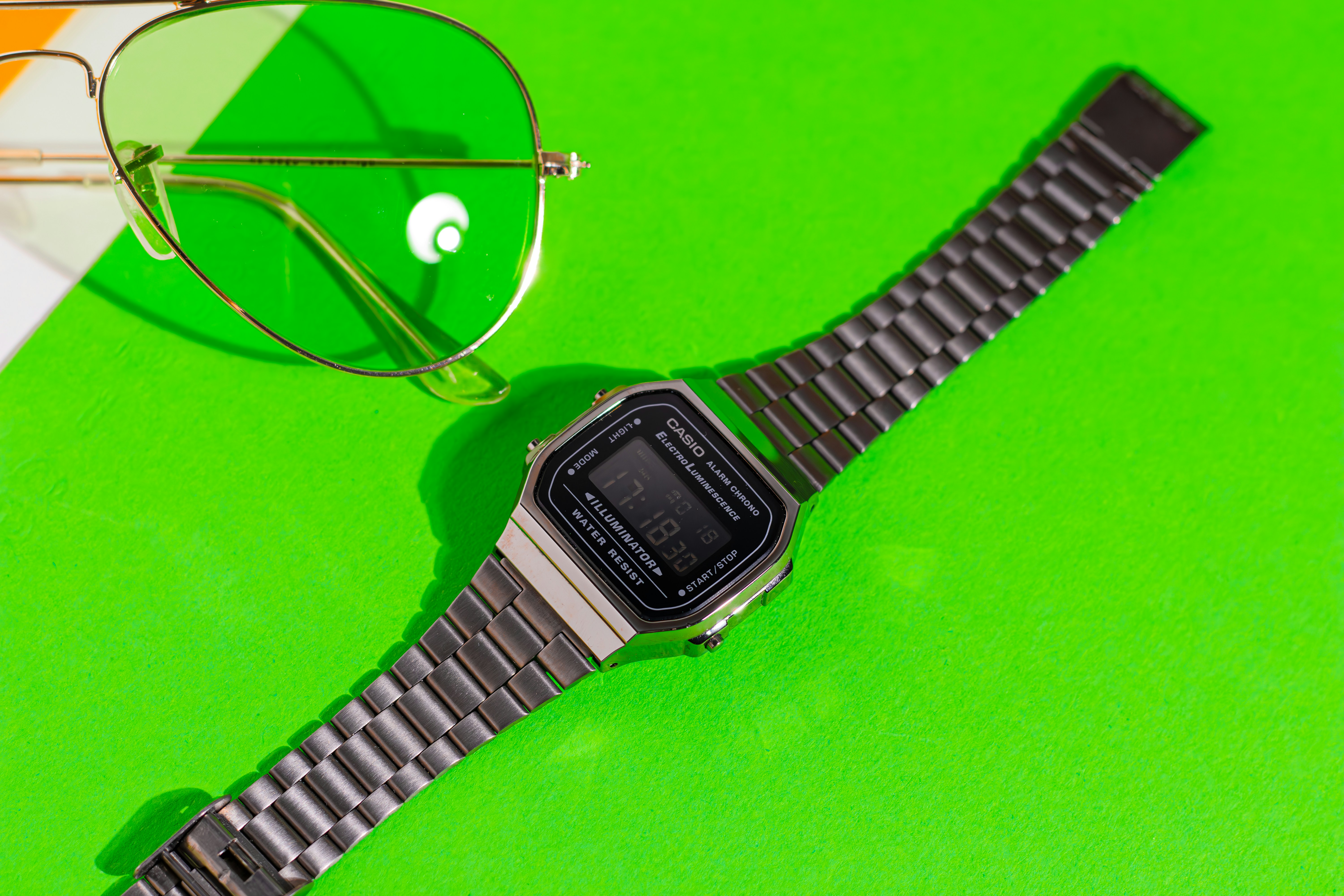 A sleek digital watch rests on vibrant green background next to stylish sunglasses, showcasing a blend of retro and contemporary fashion elements.
