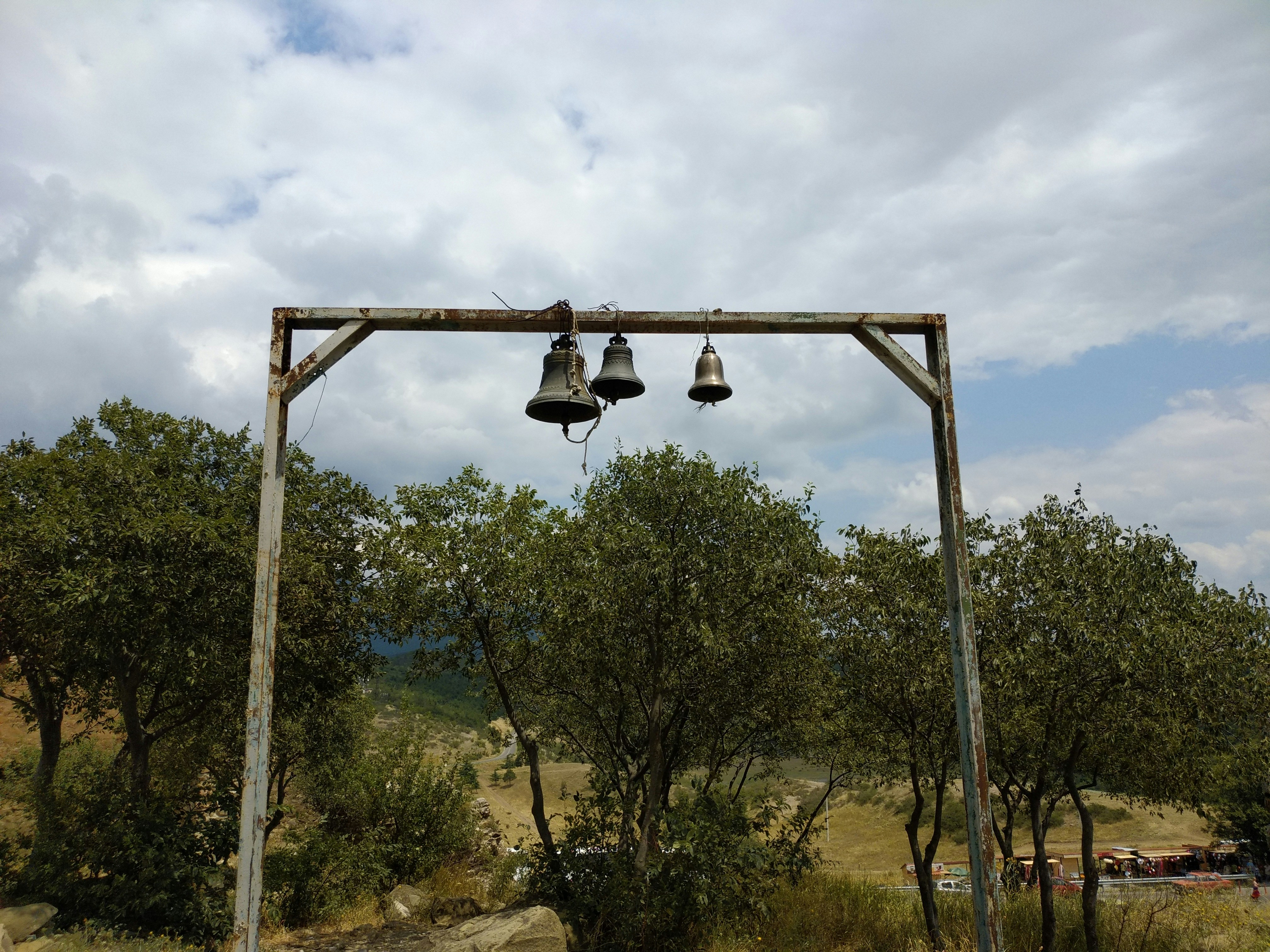 Rustic wooden frame suspends three bells against a backdrop of trees and a cloudy sky in a rural field.