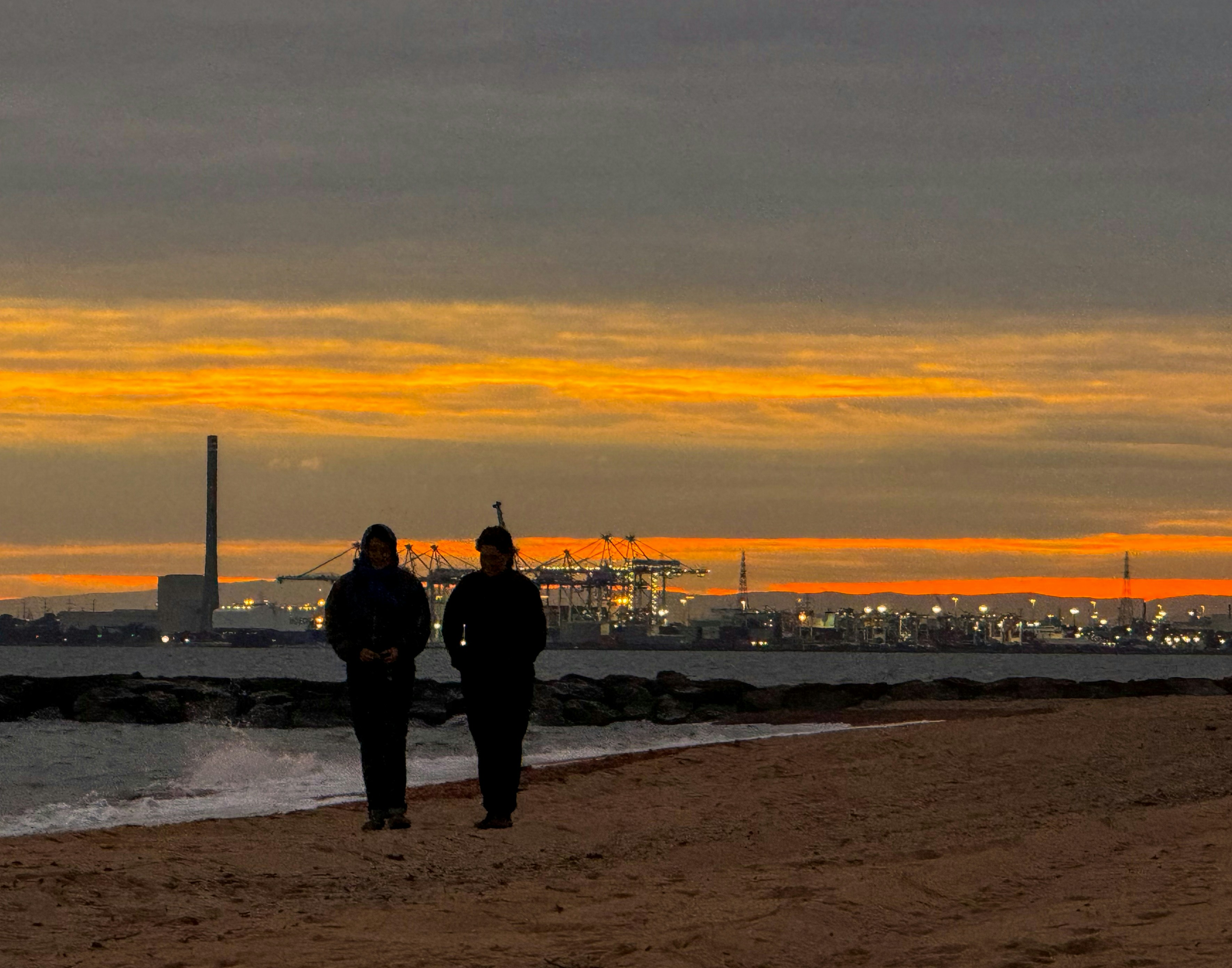 Shadows on the Shore: Two Figures Against an Industrial Dusk