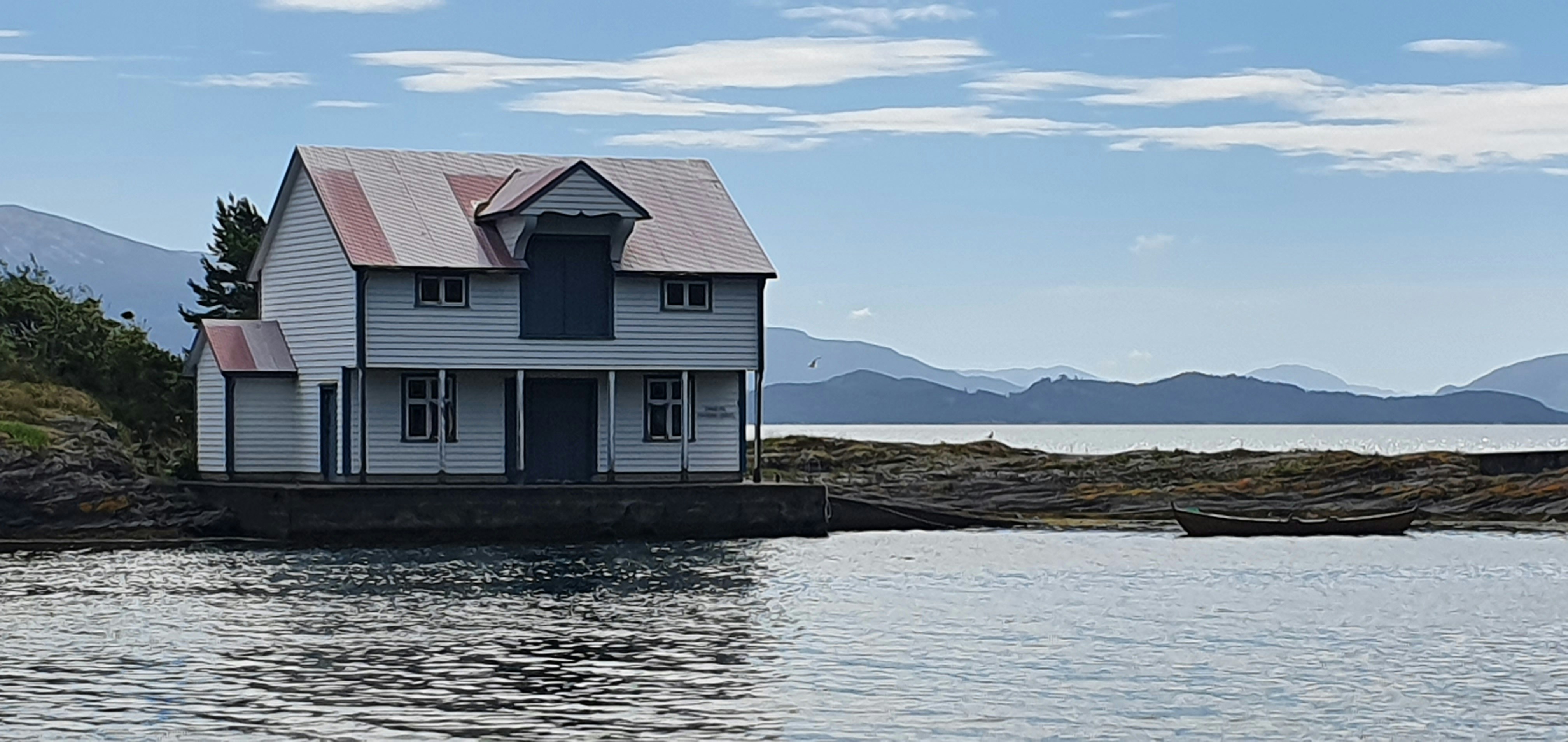 White Seaside Cottage on a Rocky Outcrop, Framed by Calm Water and Distant Peaks