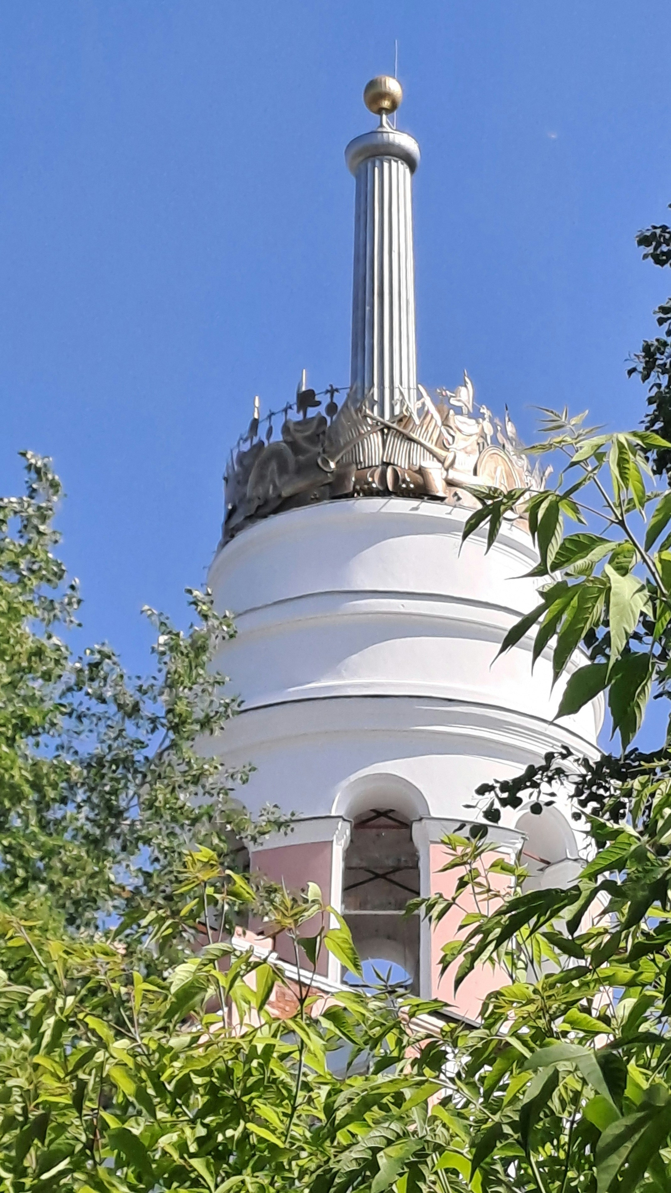 White domed tower with an ornate gilded crown rises above lush green foliage under a clear blue sky.