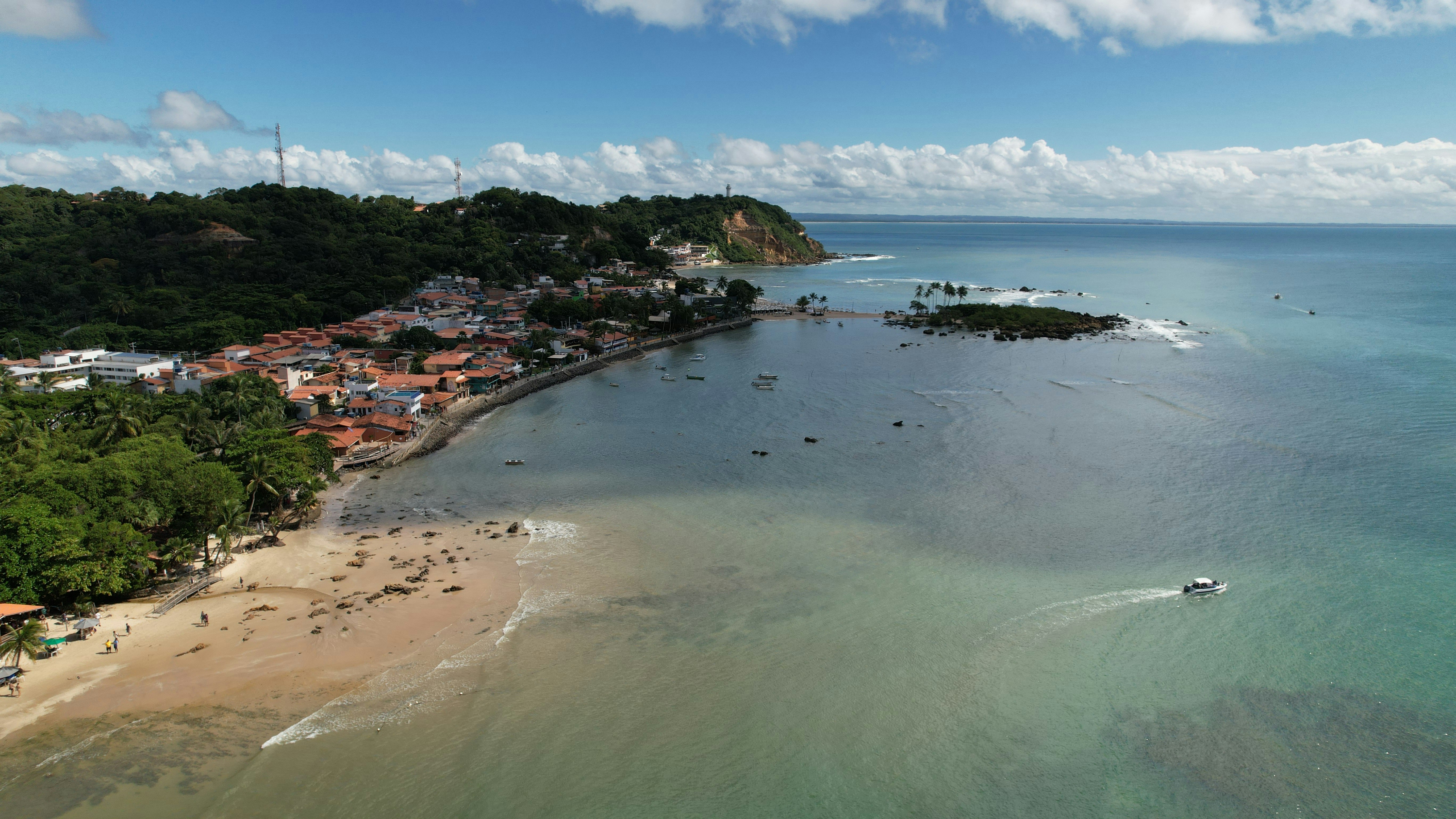 Aerial view of a coastal village with lush greenery and a clear ocean under a sky dotted with clouds.