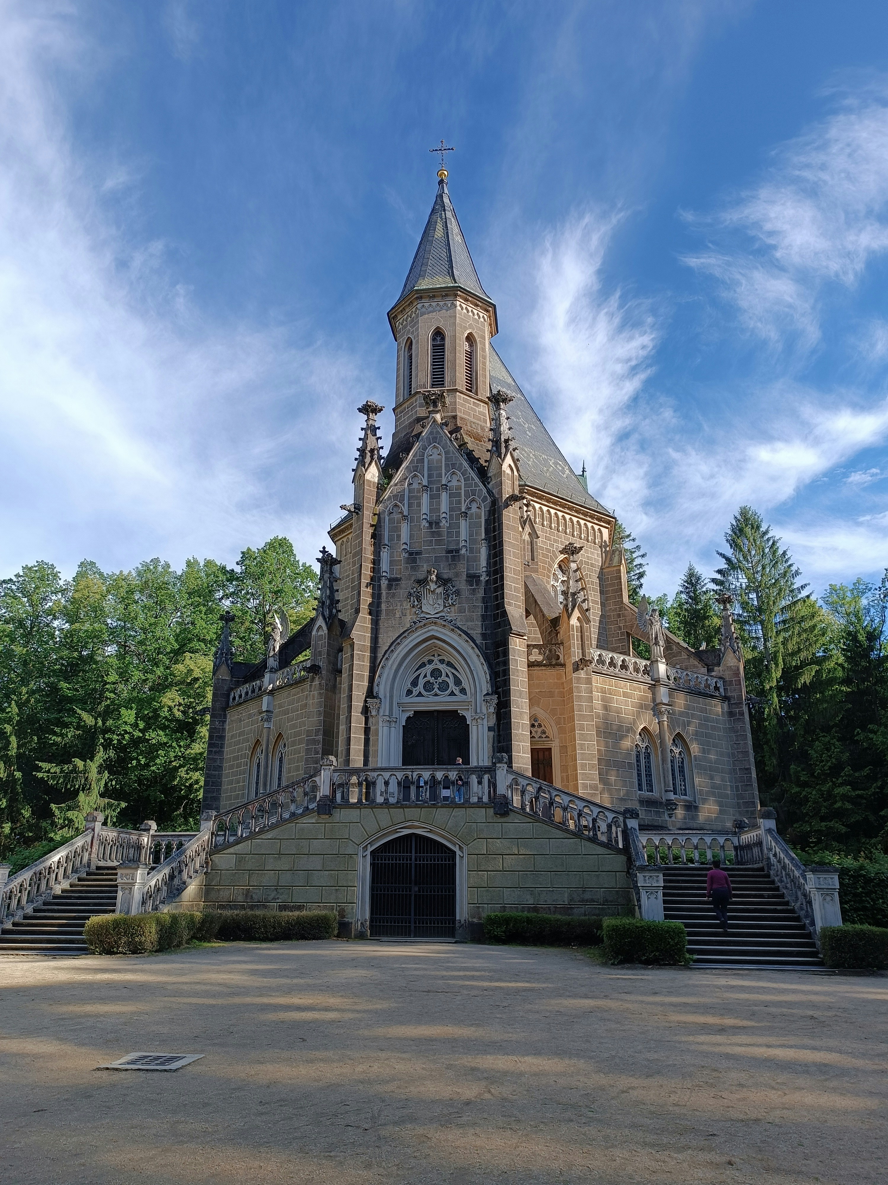 Gothic church on a terraced rise with ornate stone façade, central arched doorway, and twin staircases, framed by trees under a clear blue sky.