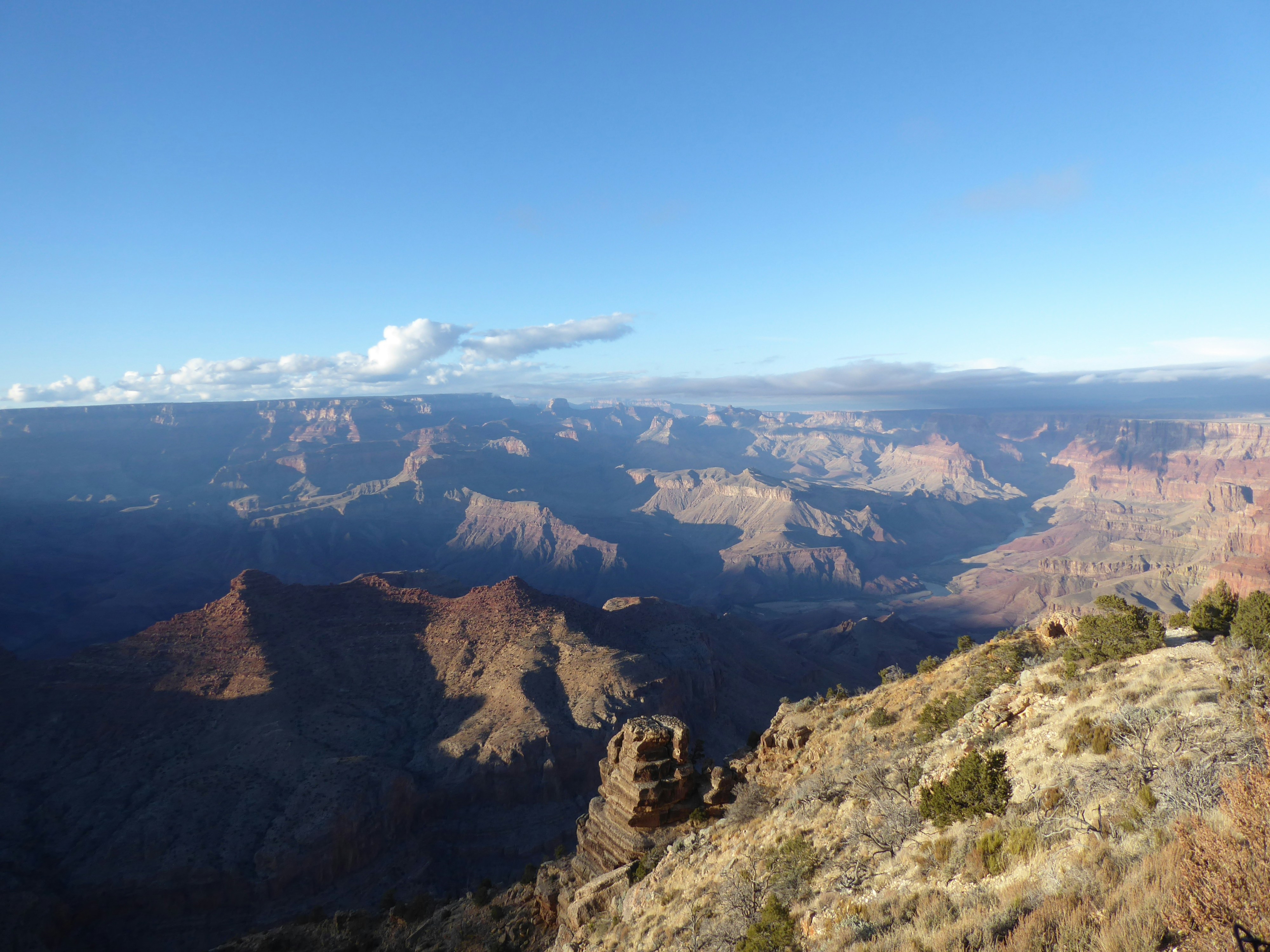 Wide landscape photograph of layered canyon cliffs beneath a clear blue sky, with sunlit foreground rocks and sparse vegetation.