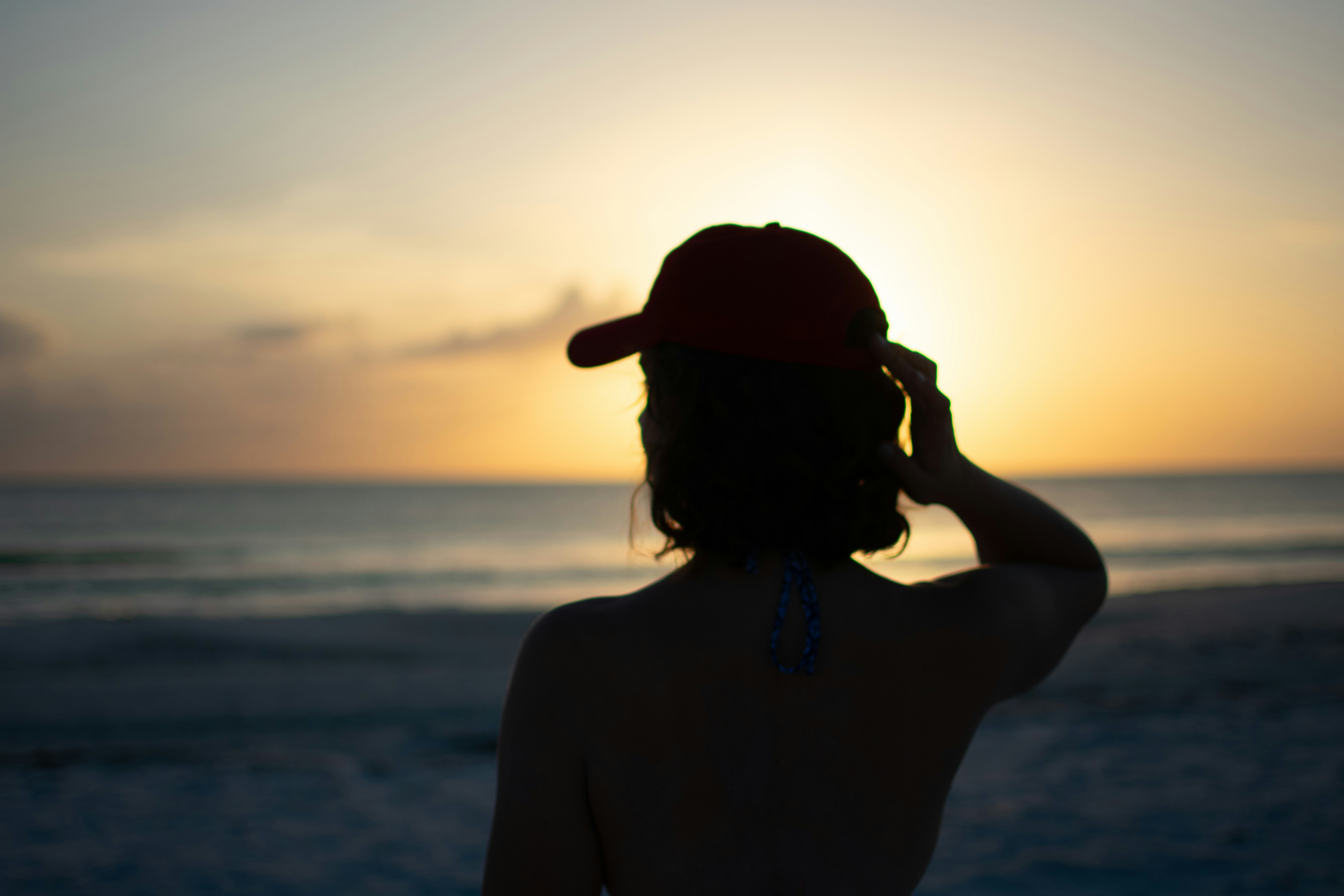 This captivating image captures a silhouette of a person wearing a red cap, gazing towards a serene ocean sunset. The composition is beautifully balanced, with the silhouette set against the warm hues of the setting sun that blend into a gradient of soft yellows and oranges. The gentle lighting and tranquil atmosphere create an evocative scene, highlighting the striking contrast between the dark figure and the radiant sky.