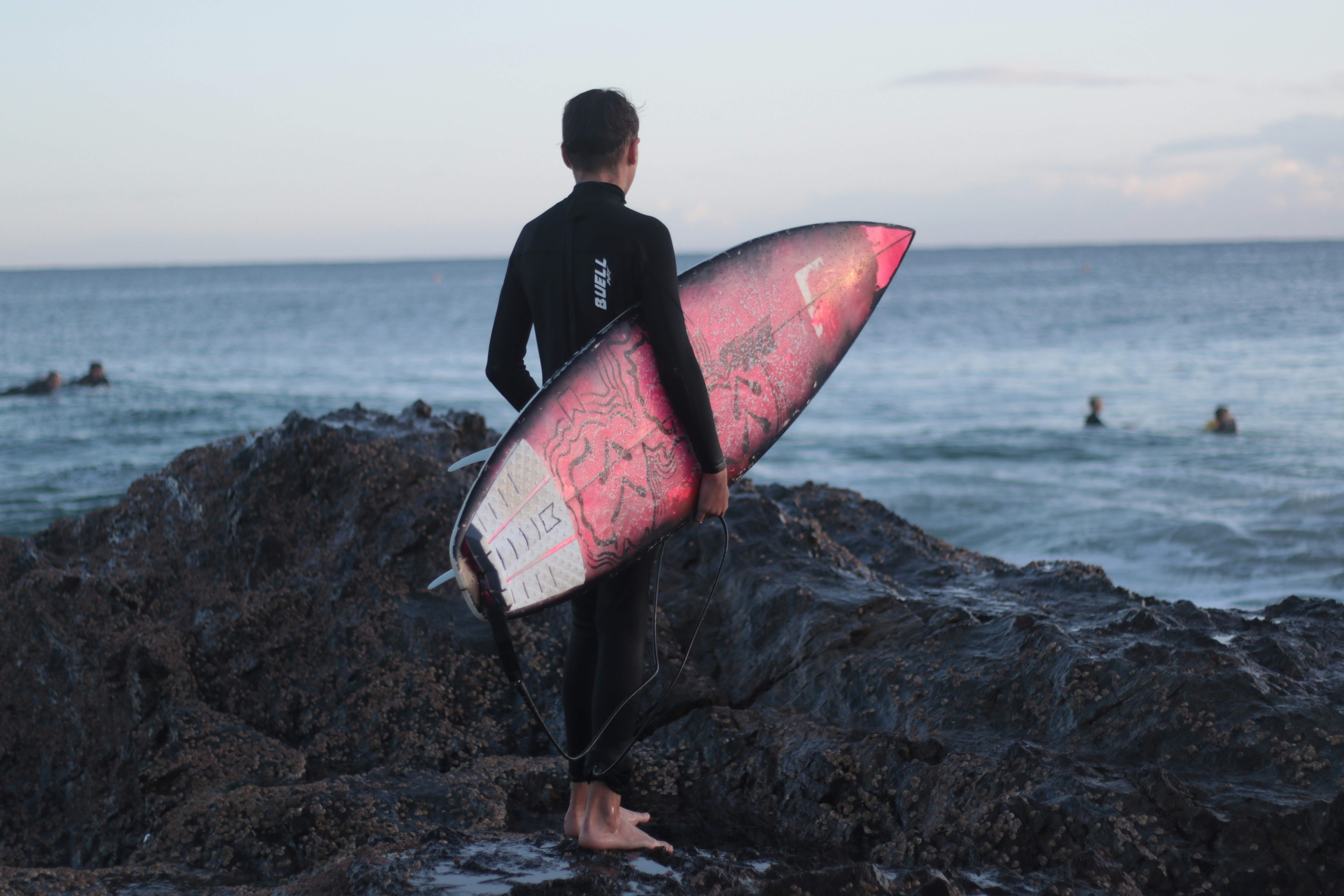 Crimson Board on a Quiet Edge: Surfer Pauses on Lava Rocks at Dusk