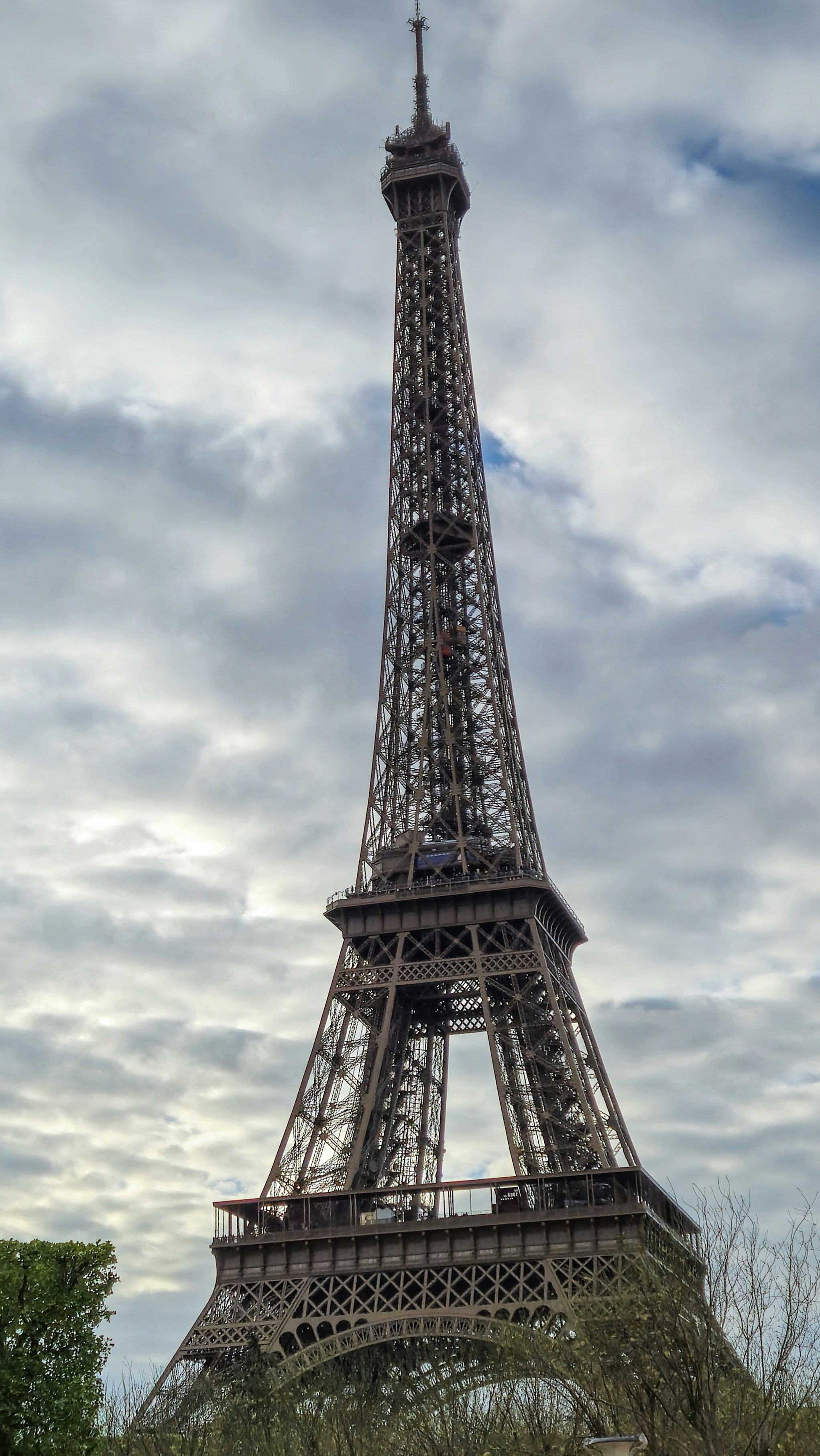 Eiffel Tower's iron lattice rises against a cloudy sky, with greenery visible at the base.