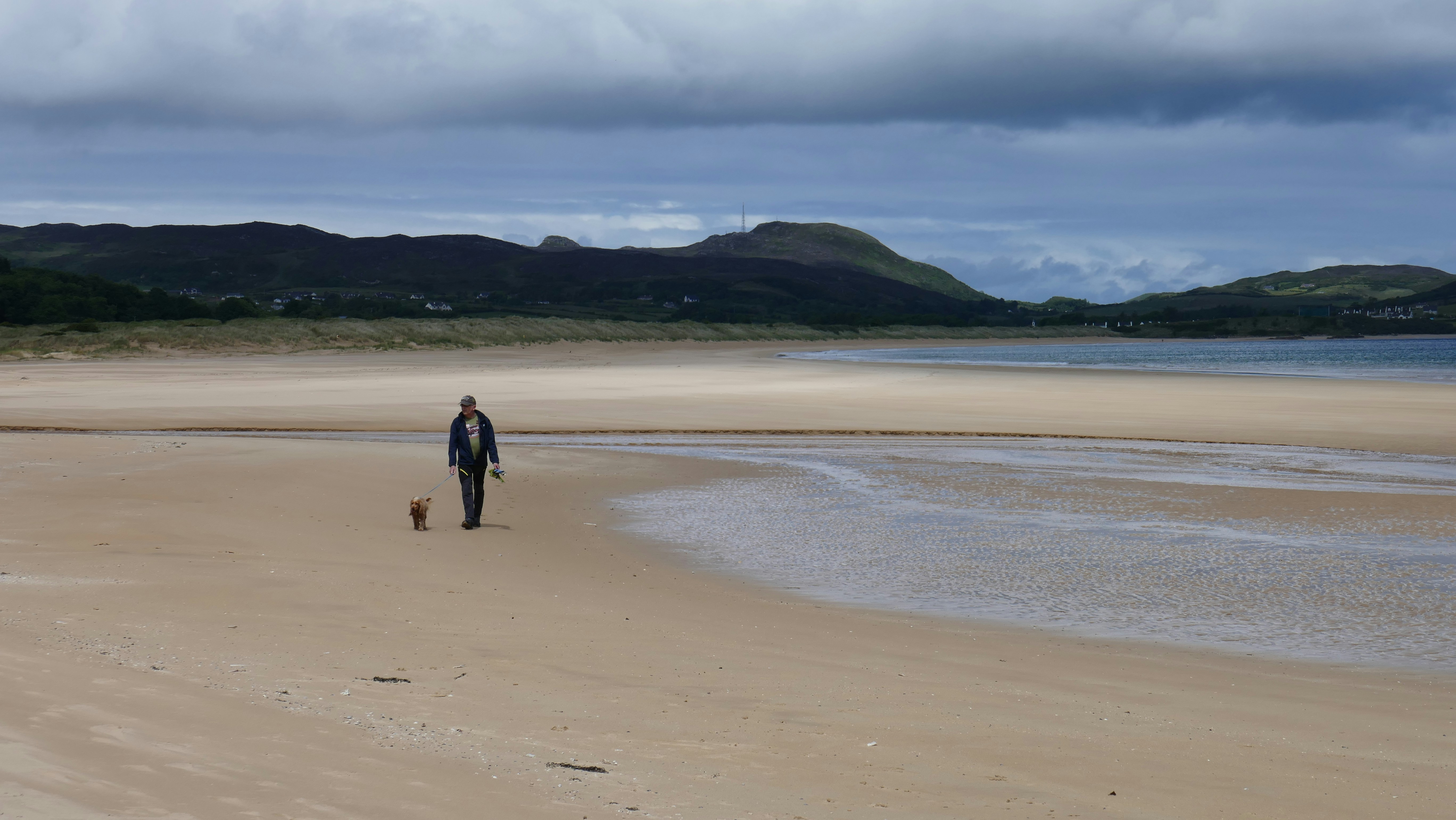 A solitary figure walks a small dog along a broad, wind-swept beach, with a shallow tidal pool curling toward the shore. Distant hills under a moody sky provide a dramatic backdrop.