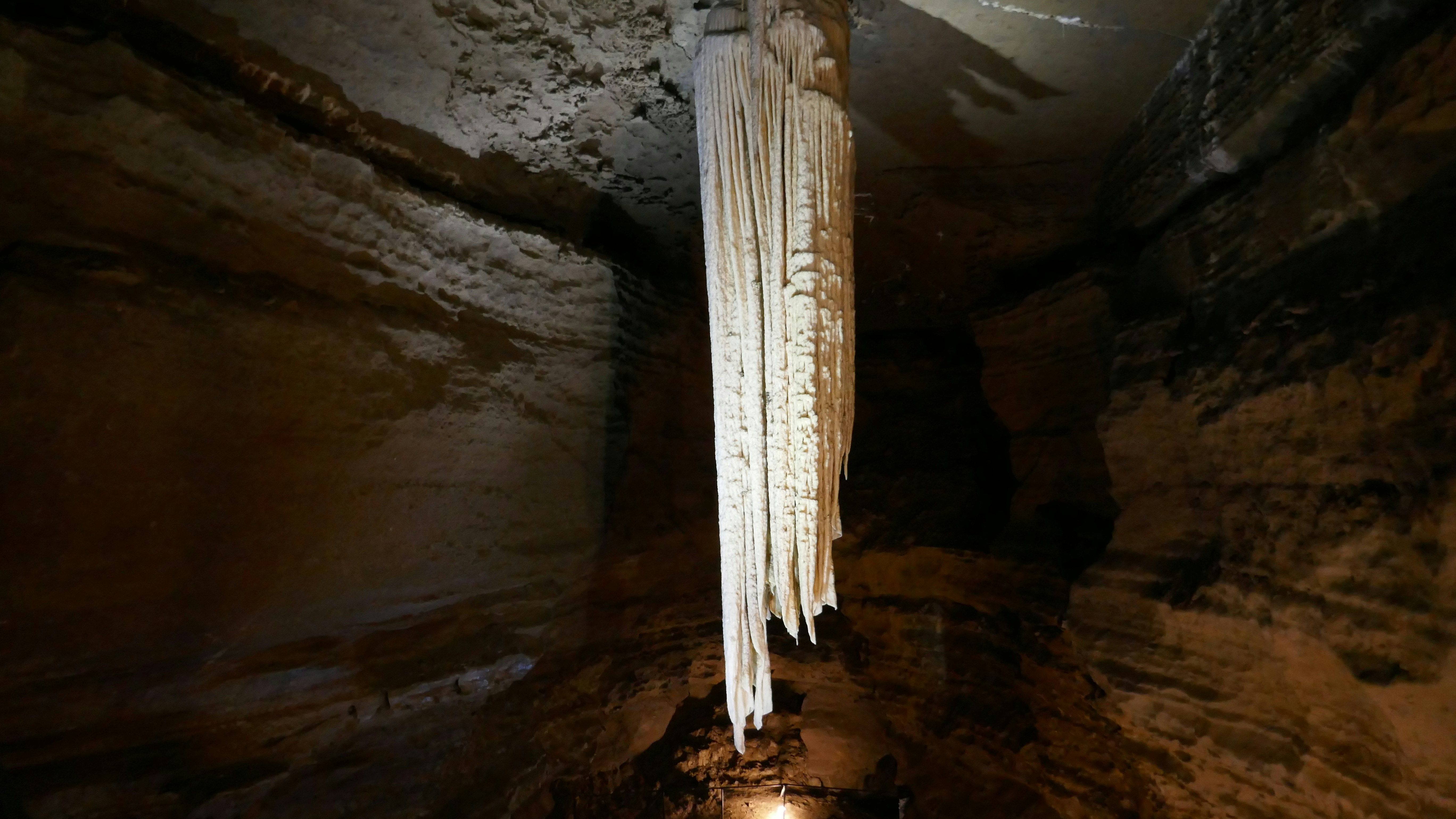 A photograph of a narrow limestone cavern with a solitary hanging stalactite against rugged rock walls.