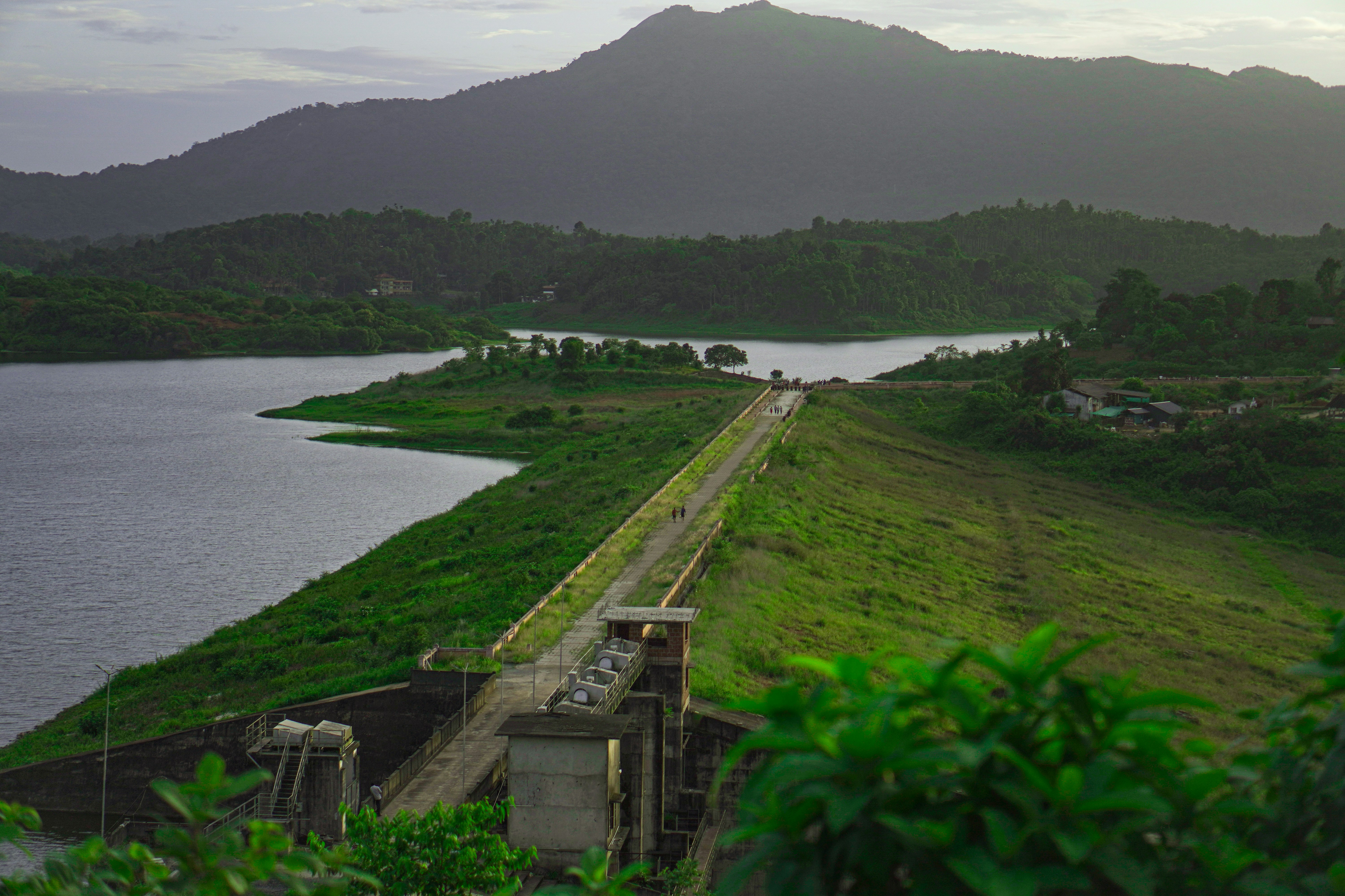 A serene dam road winding through lush greenery beside a calm lake, framed by distant mountains. The landscape reflects harmony between human engineering and natural beauty.