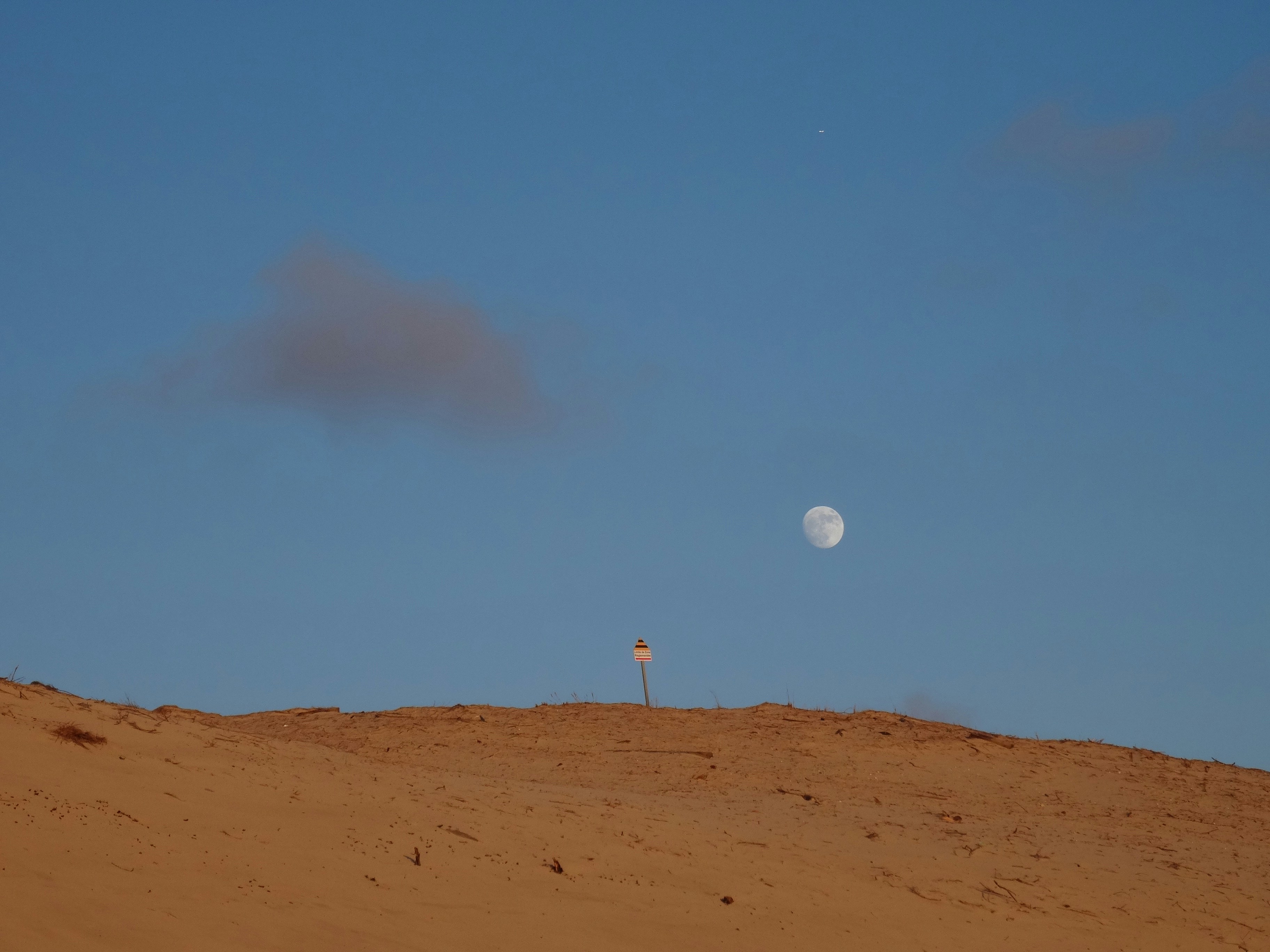 A lone signpost sits atop a sandy dune beneath a clear blue sky. This photograph highlights a visible moon near the horizon.