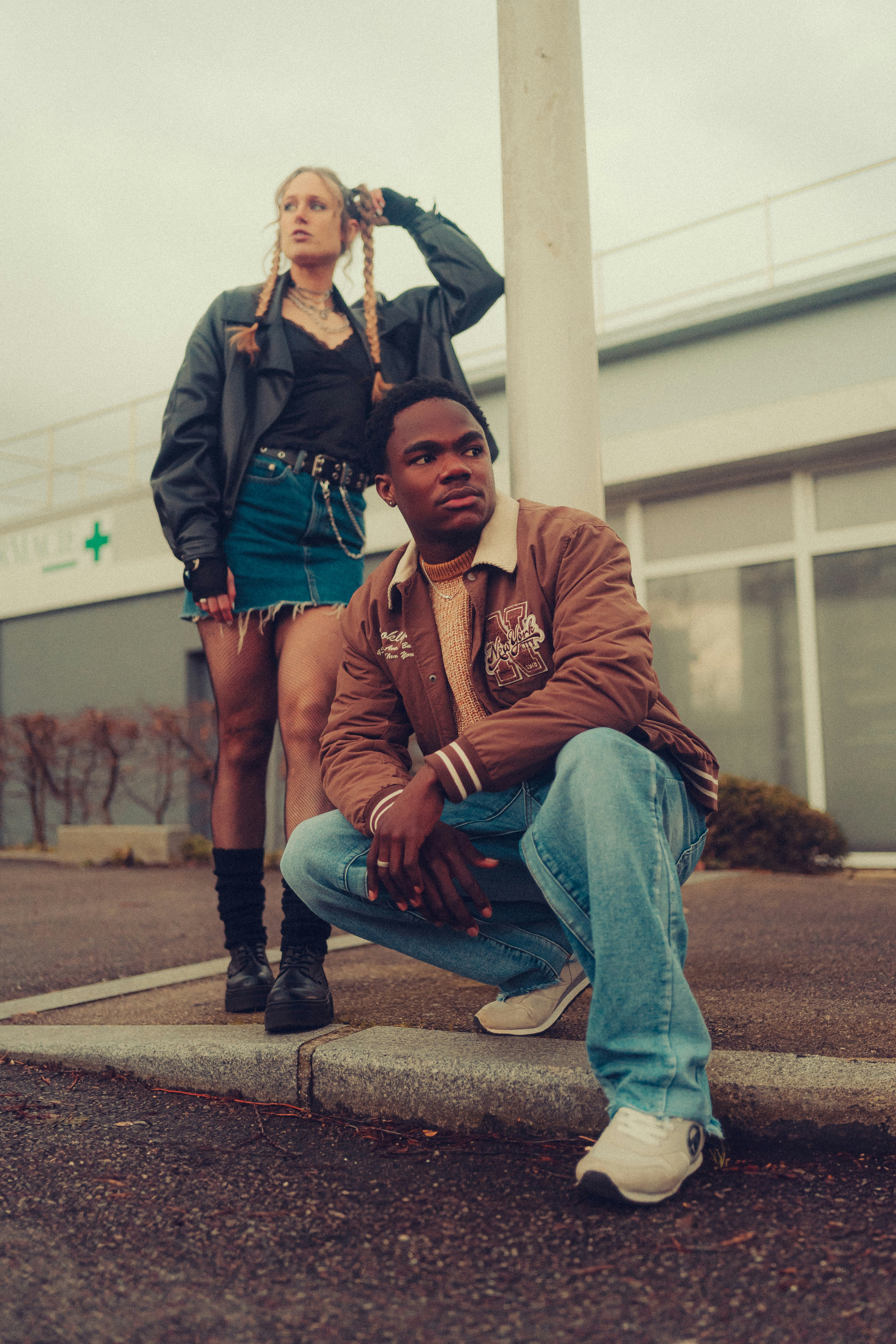 Two stylish individuals pose in an urban setting, showcasing contemporary fashion against a muted backdrop. The scene captures a blend of confidence and casual elegance.