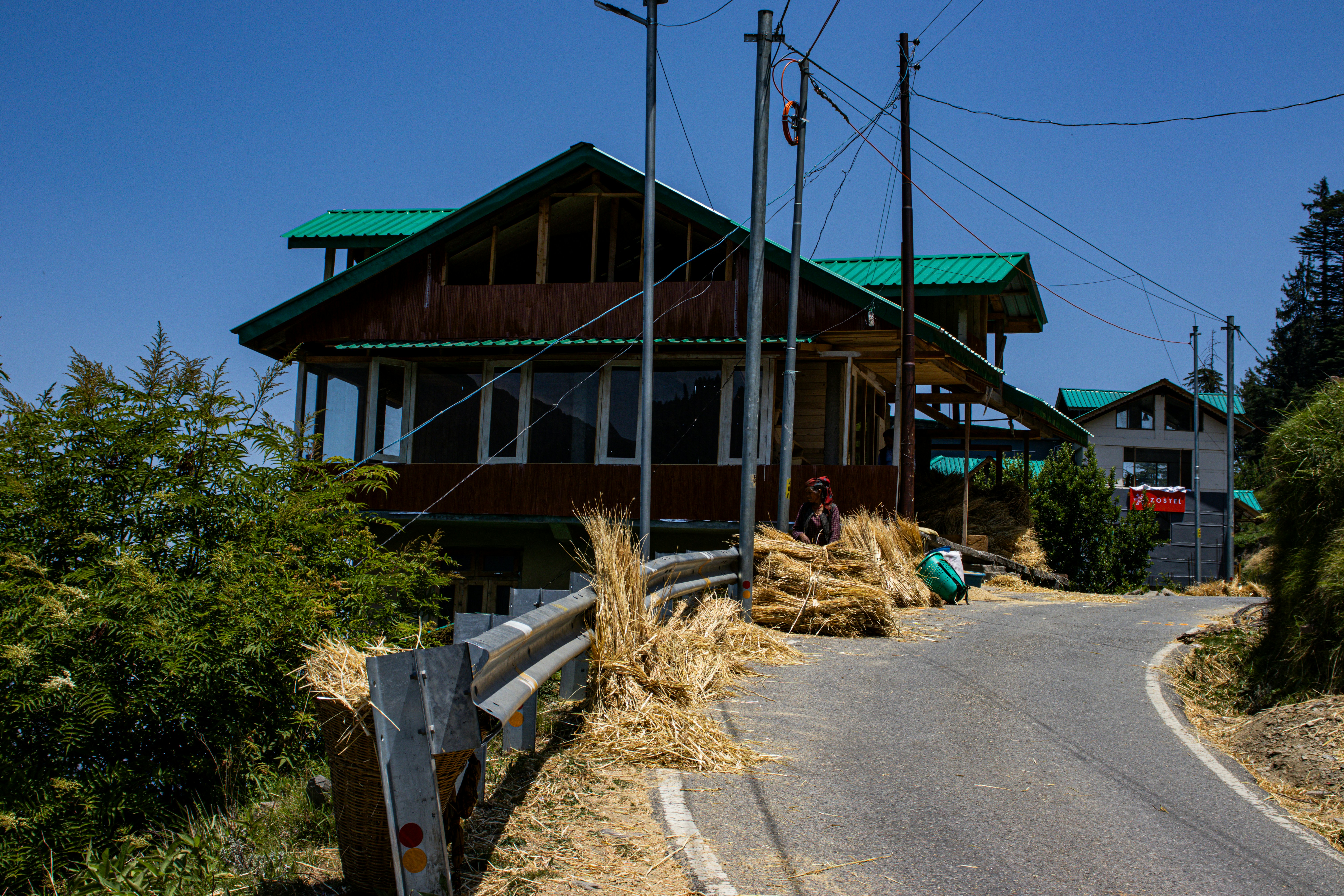 A rural scene showcasing a winding road bordered by a wooden house and workers gathering straw under a clear blue sky.