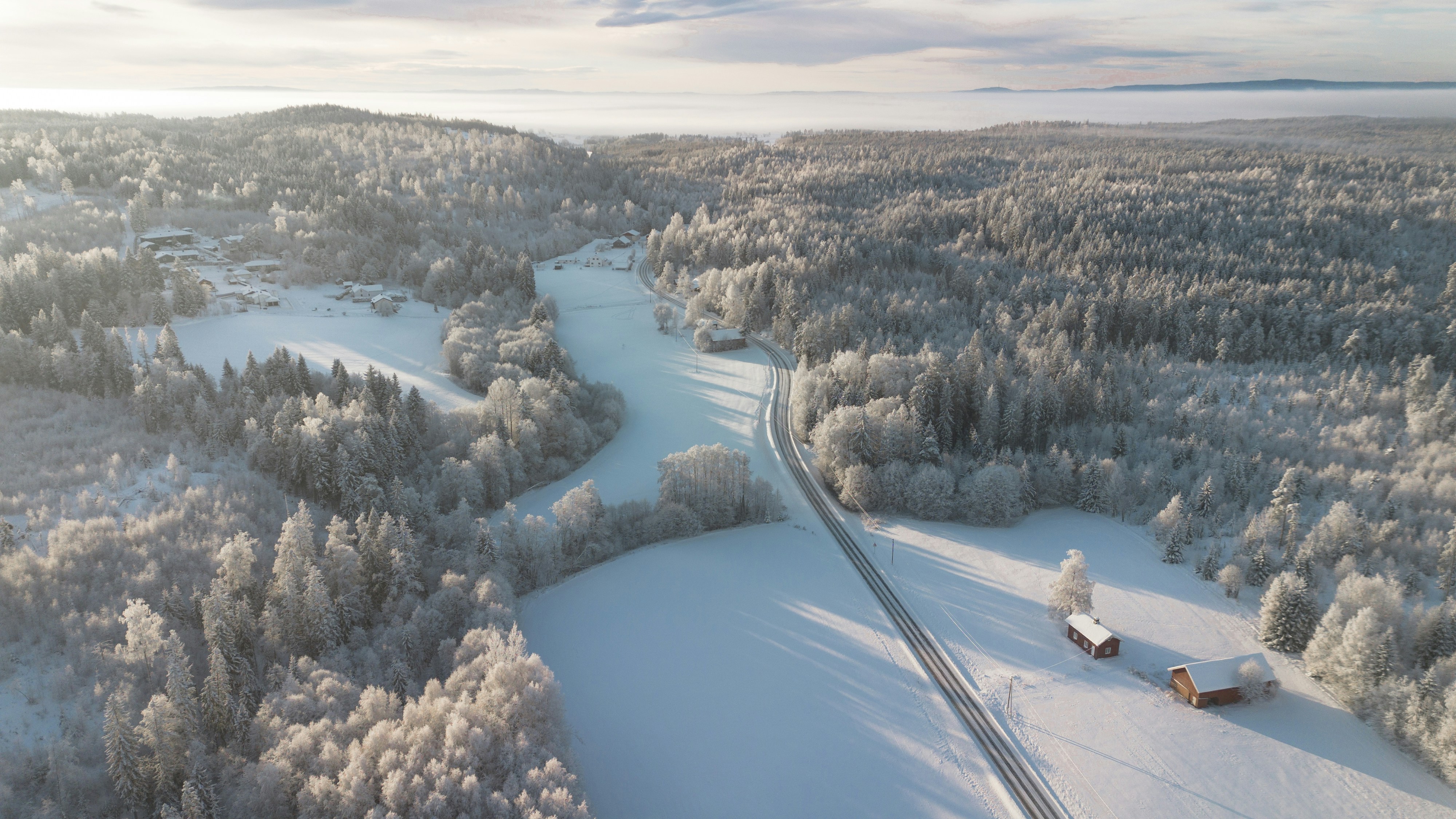 Aerial drone photo of a snow-blanketed forest with a winding river cutting through the valley and a pair of wooden cabins in the lower-right under soft winter light.