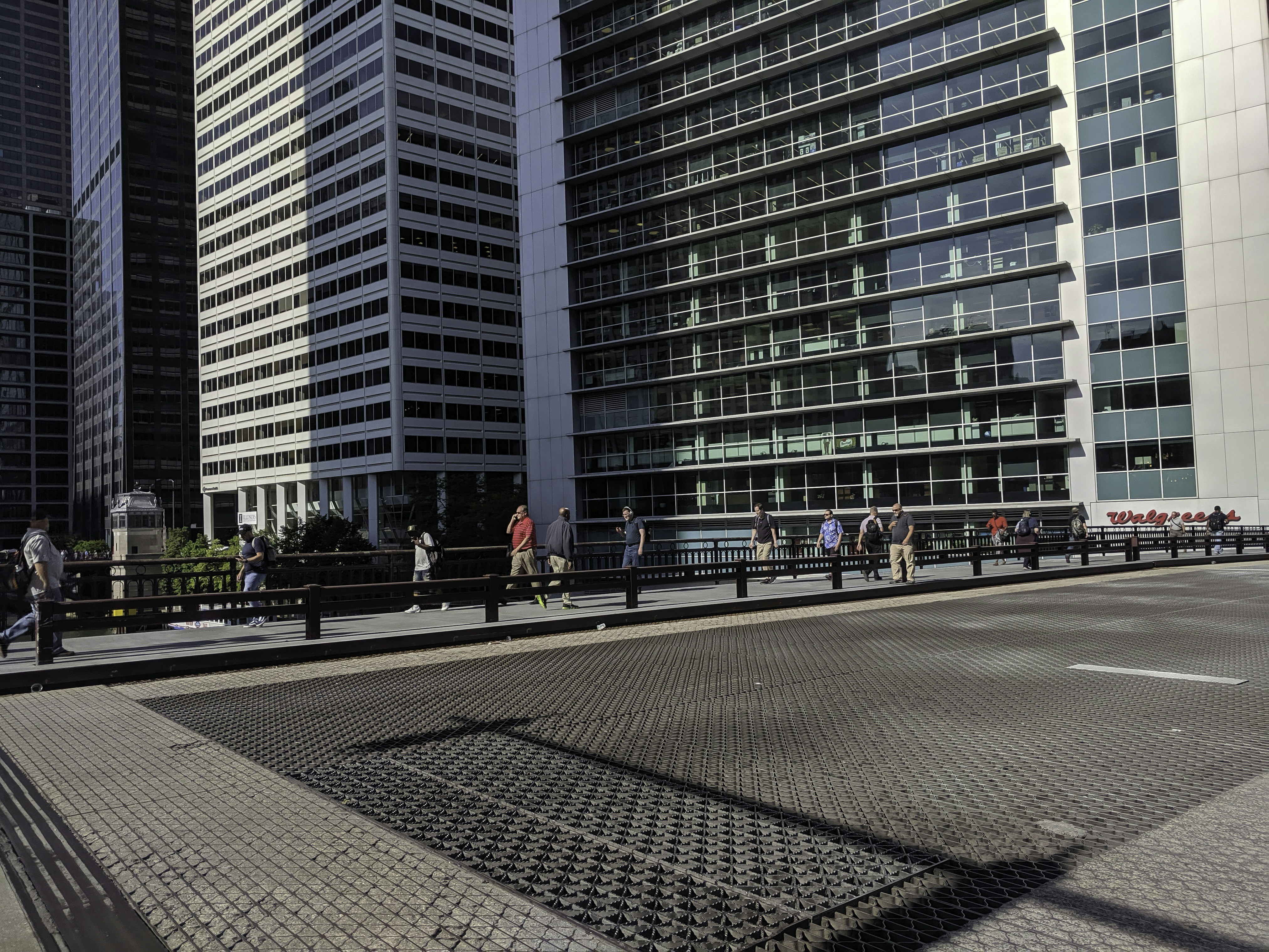 Urban street scene with pedestrians along a railing beside tall glass office buildings under daylight.