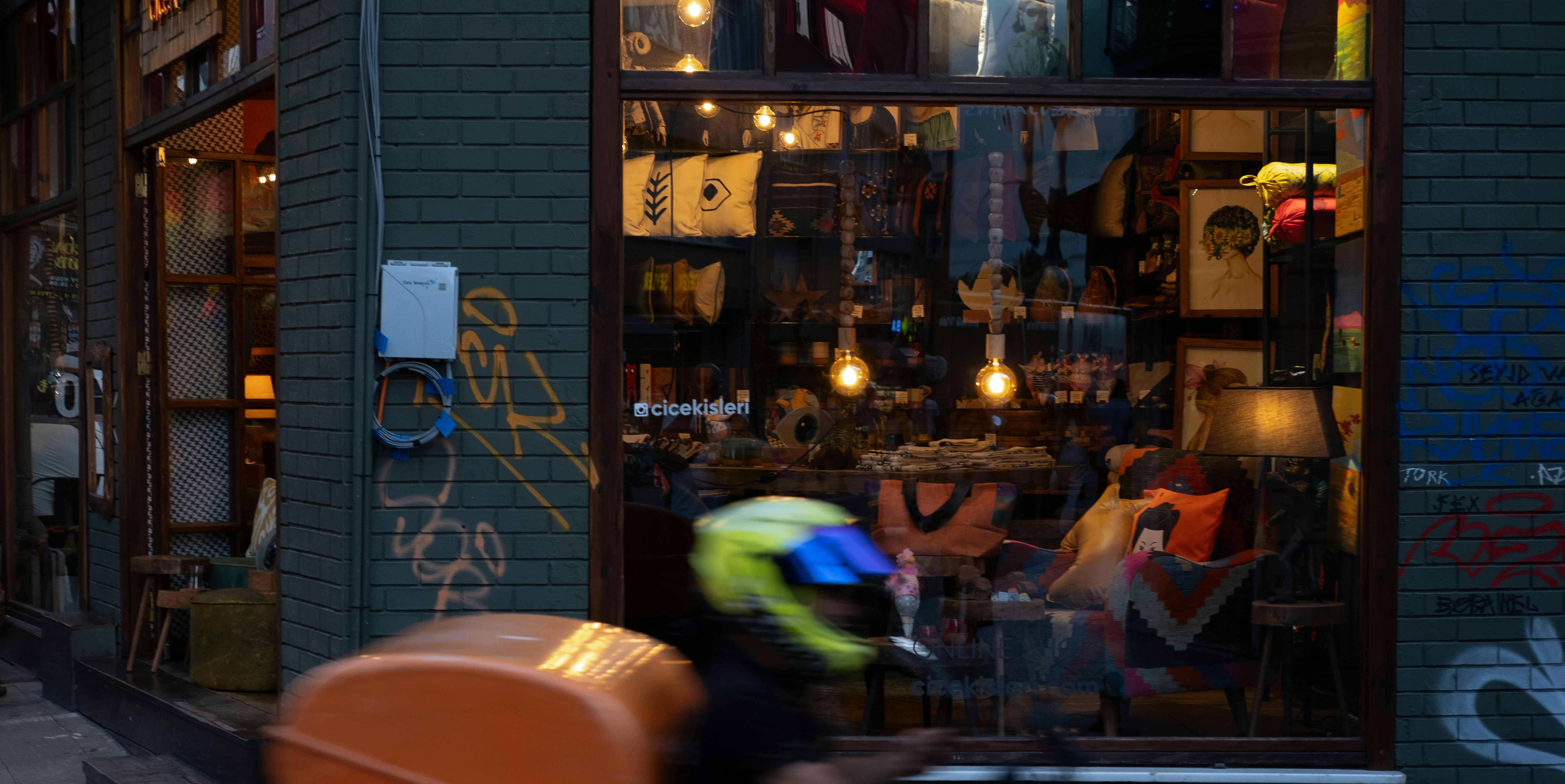 Cozy Istanbul storefront filled with colorful decor, warm interior lighting contrasting with dark, graffiti-covered exterior. Motorcyclist with yellow helmet adds dynamic movement.