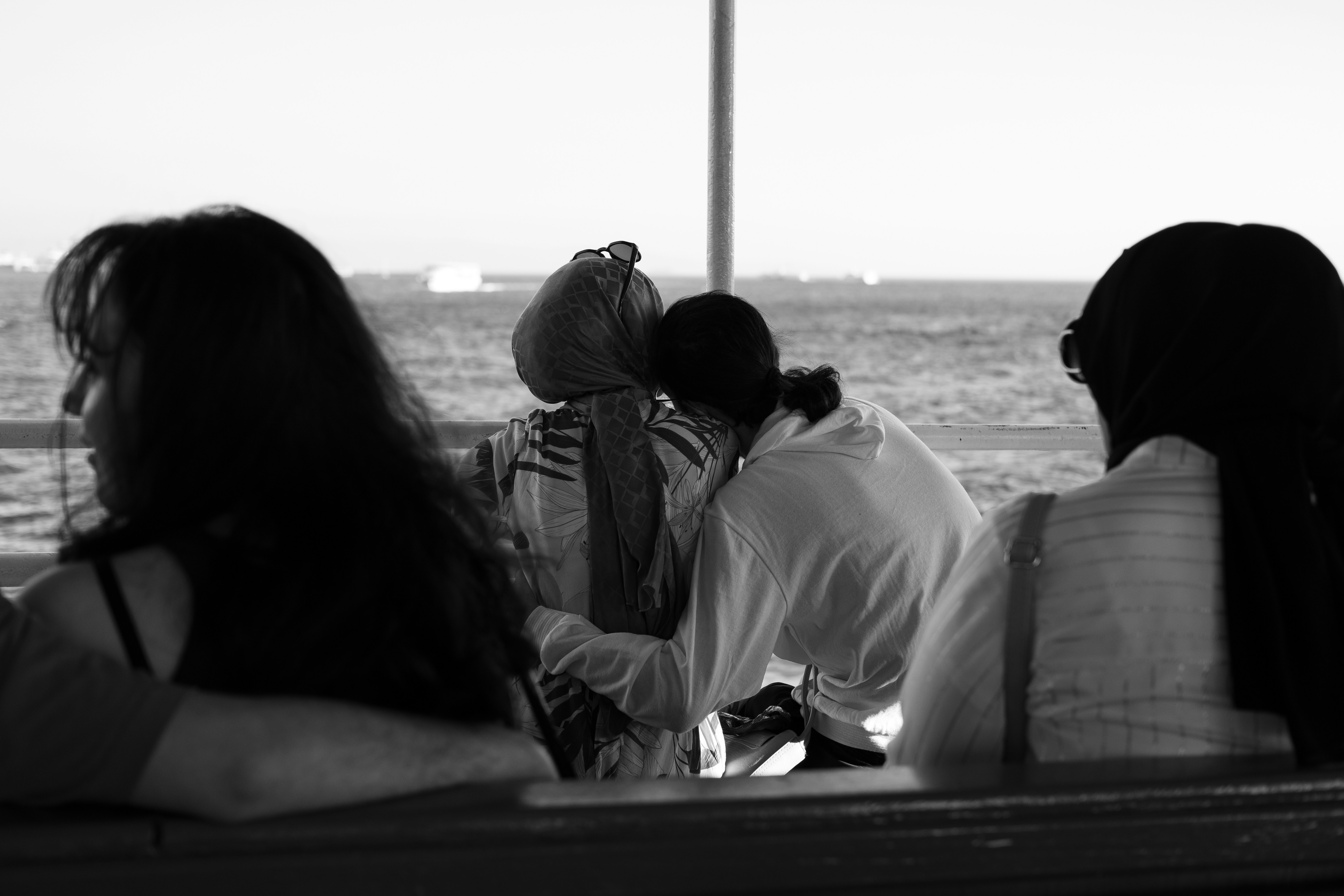 This black and white image captures a moment of serene companionship as two people sit closely on a ferry, gazing out at the open sea. The composition centers on their intimate connection, with the soft lighting highlighting the gentle folds of their clothing against the vast, textured ocean backdrop. The monochrome palette enhances the timeless and contemplative atmosphere, making the scene visually striking and emotionally resonant.