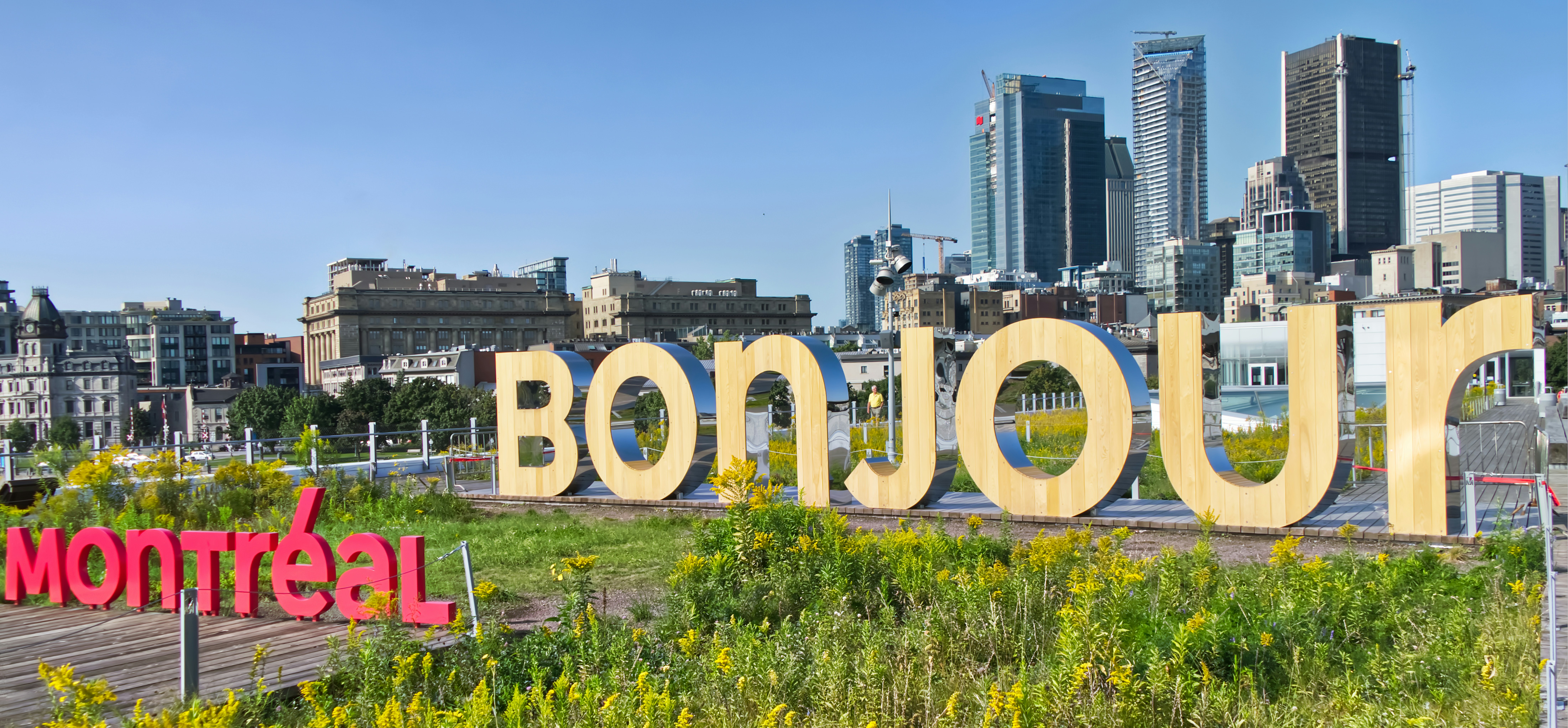 Large wooden letters spelling 'BONJOUR' in a vibrant urban park with city skyline in the background. The word 'Montréal' is displayed in bold pink, adding a pop of color.