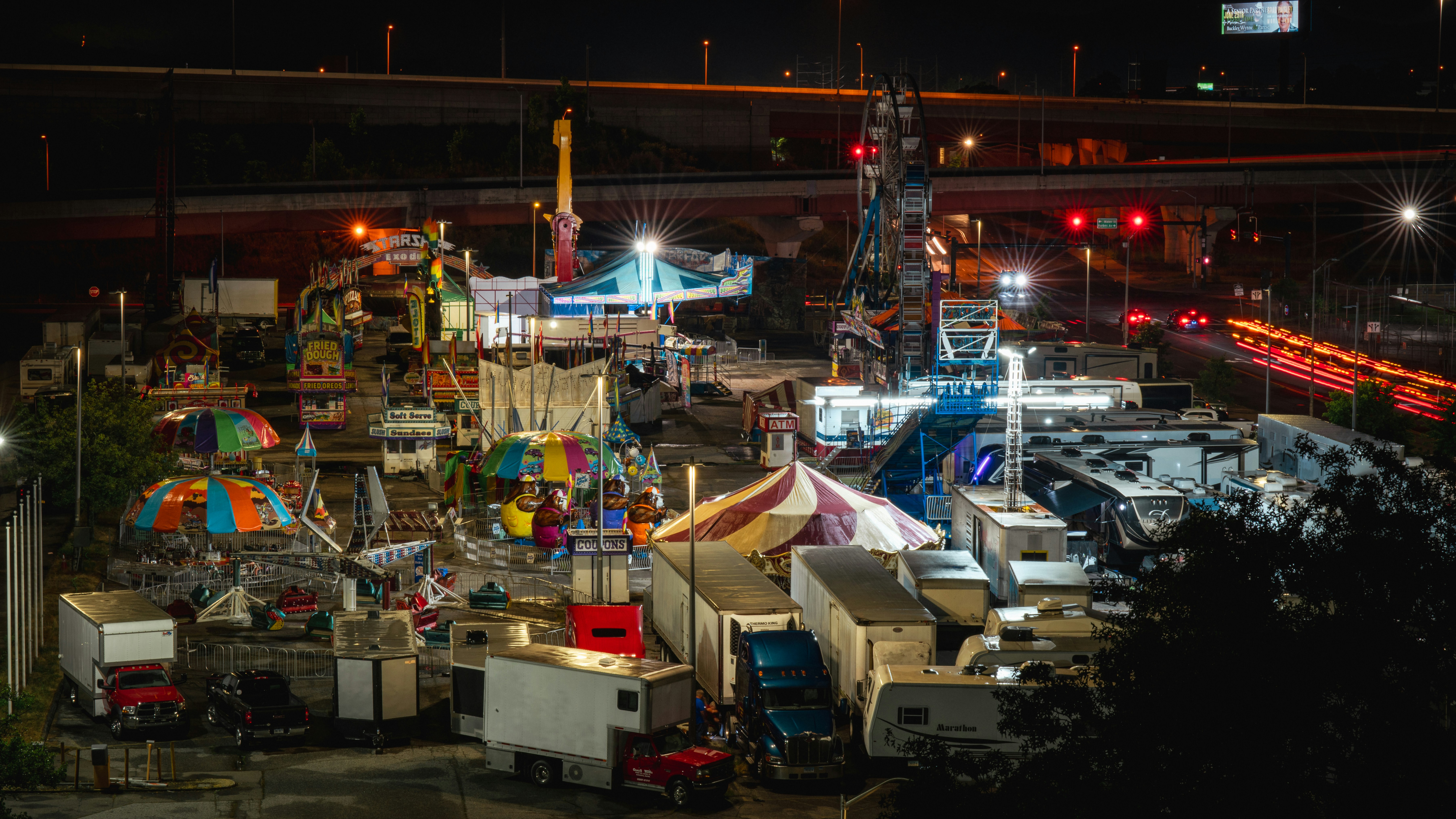 Vibrant carnival rides and booths illuminated under night lights, surrounded by parked vehicles and urban infrastructure.