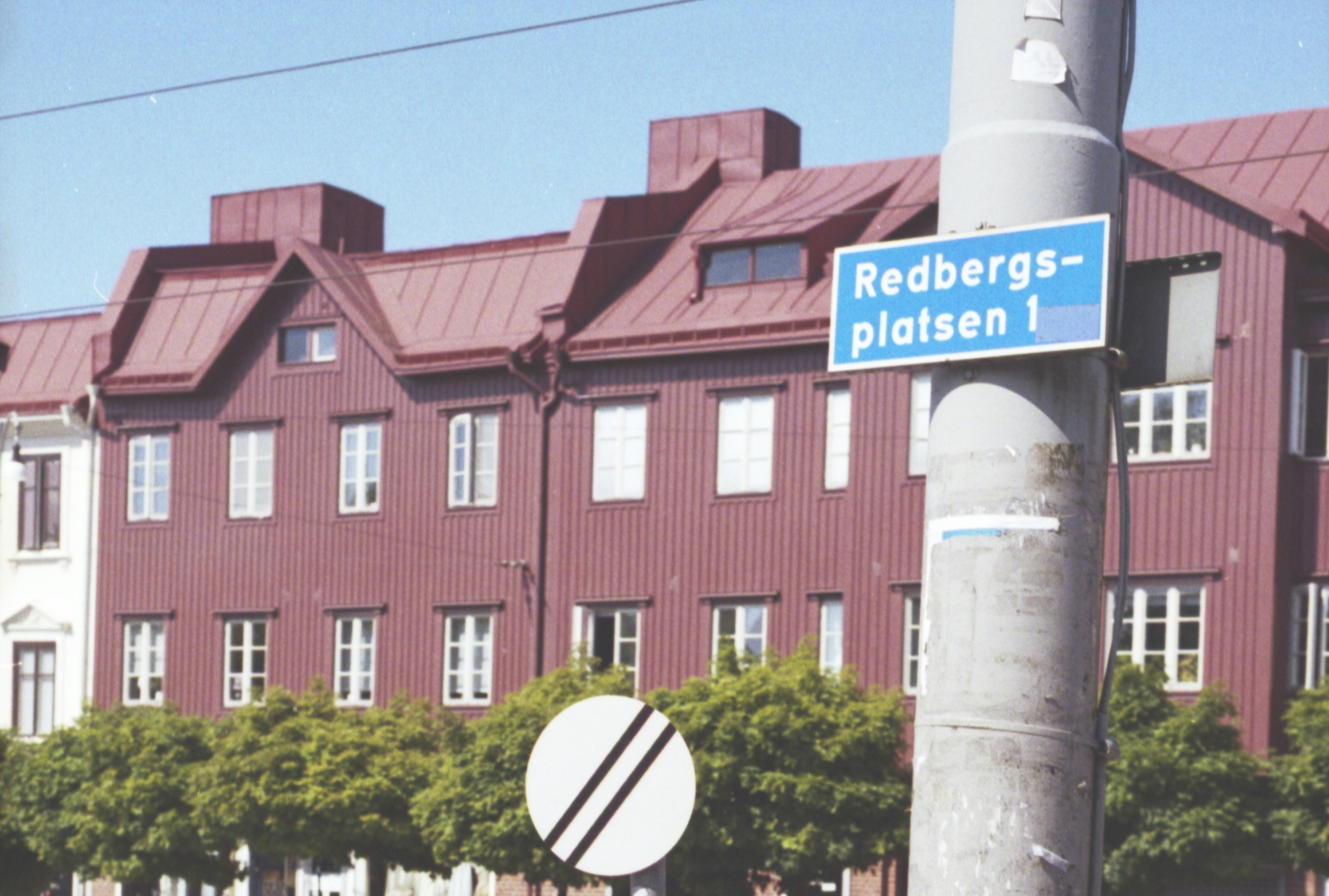 Row of red buildings with uniform windows under a clear blue sky, featuring a Redbergsplatsen street sign on a lamppost.