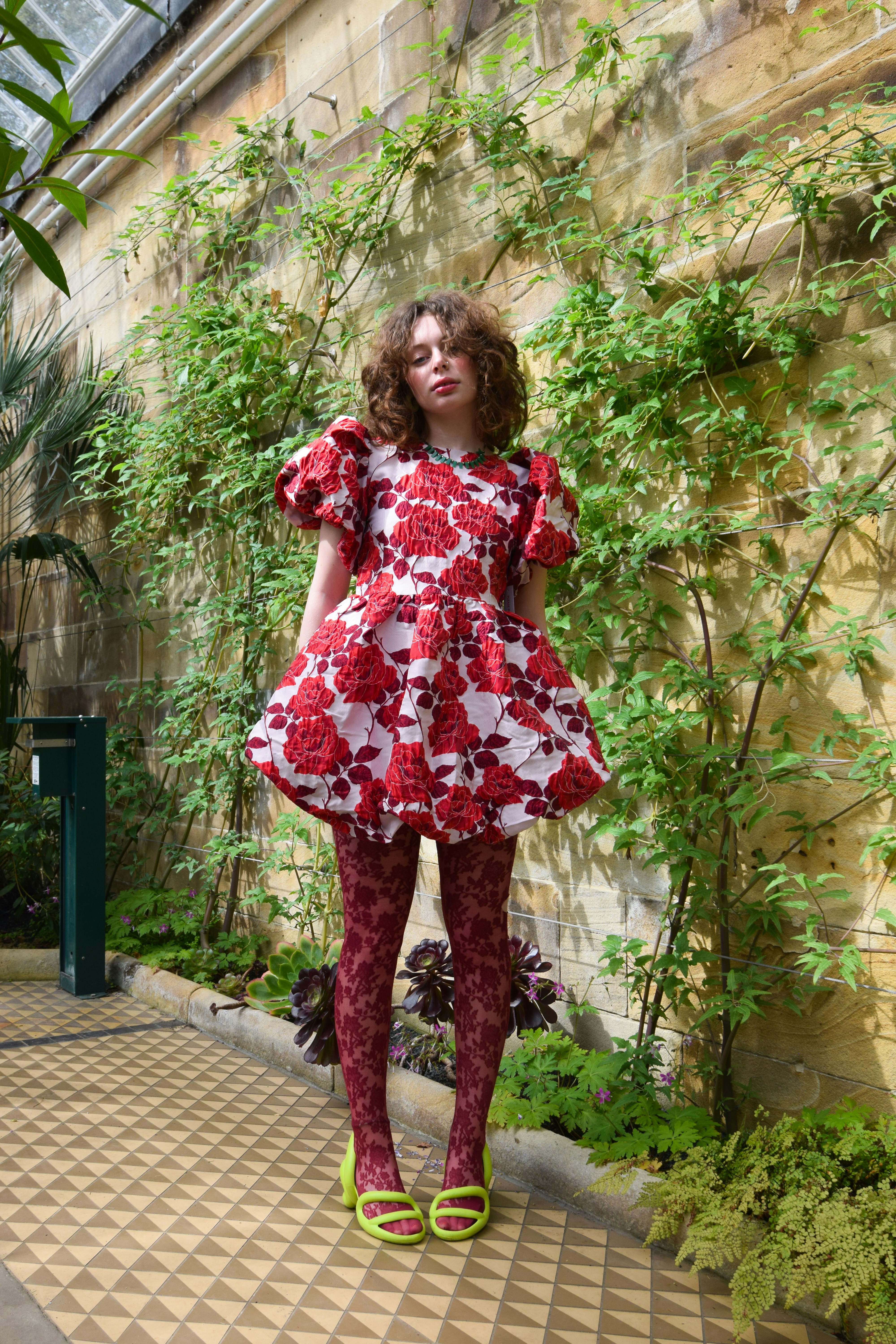 Model in a vibrant floral dress stands amidst a verdant backdrop, showcasing a unique blend of fashion and nature.