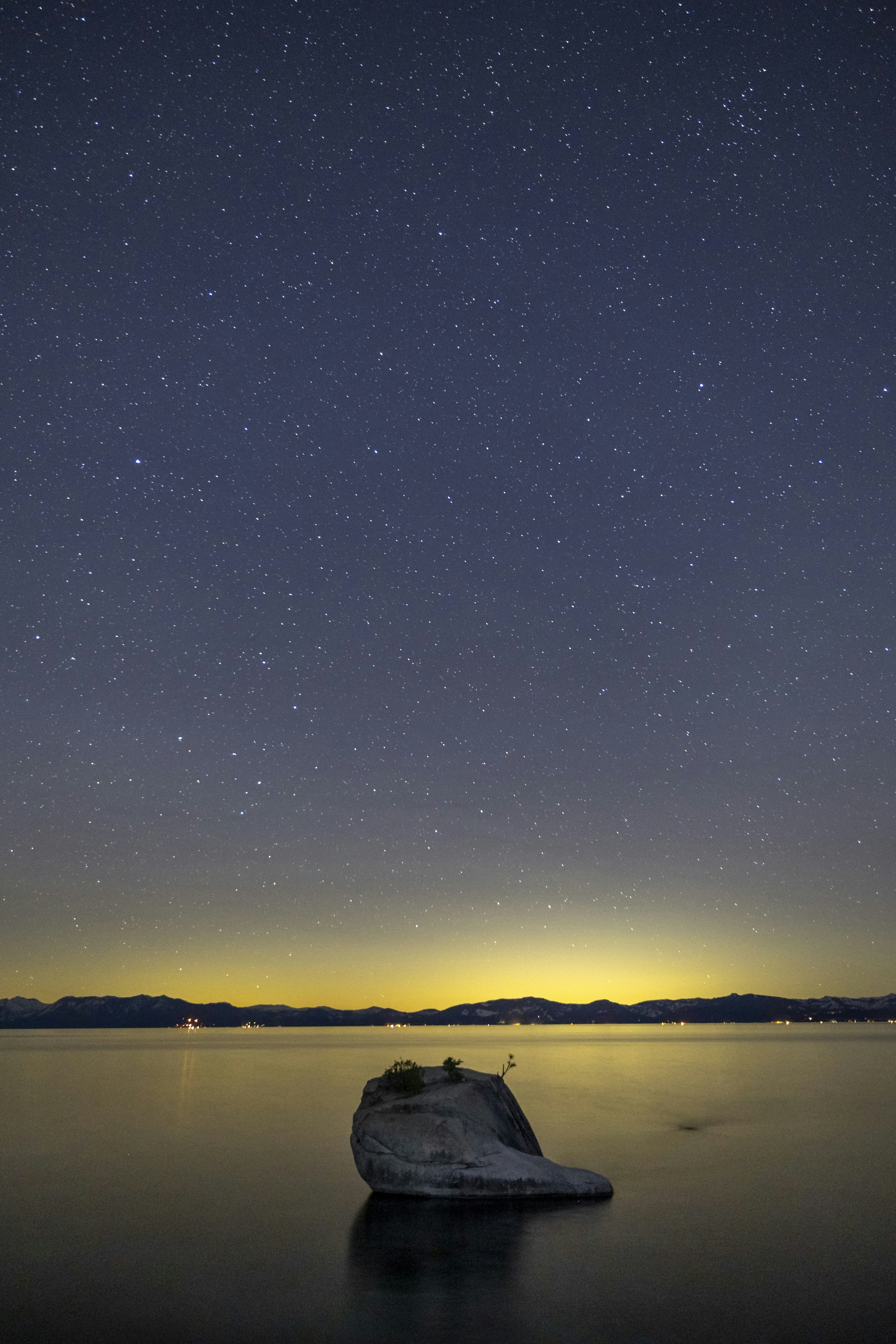 Night photograph of a star-filled sky over a calm lake with a lone rock.
