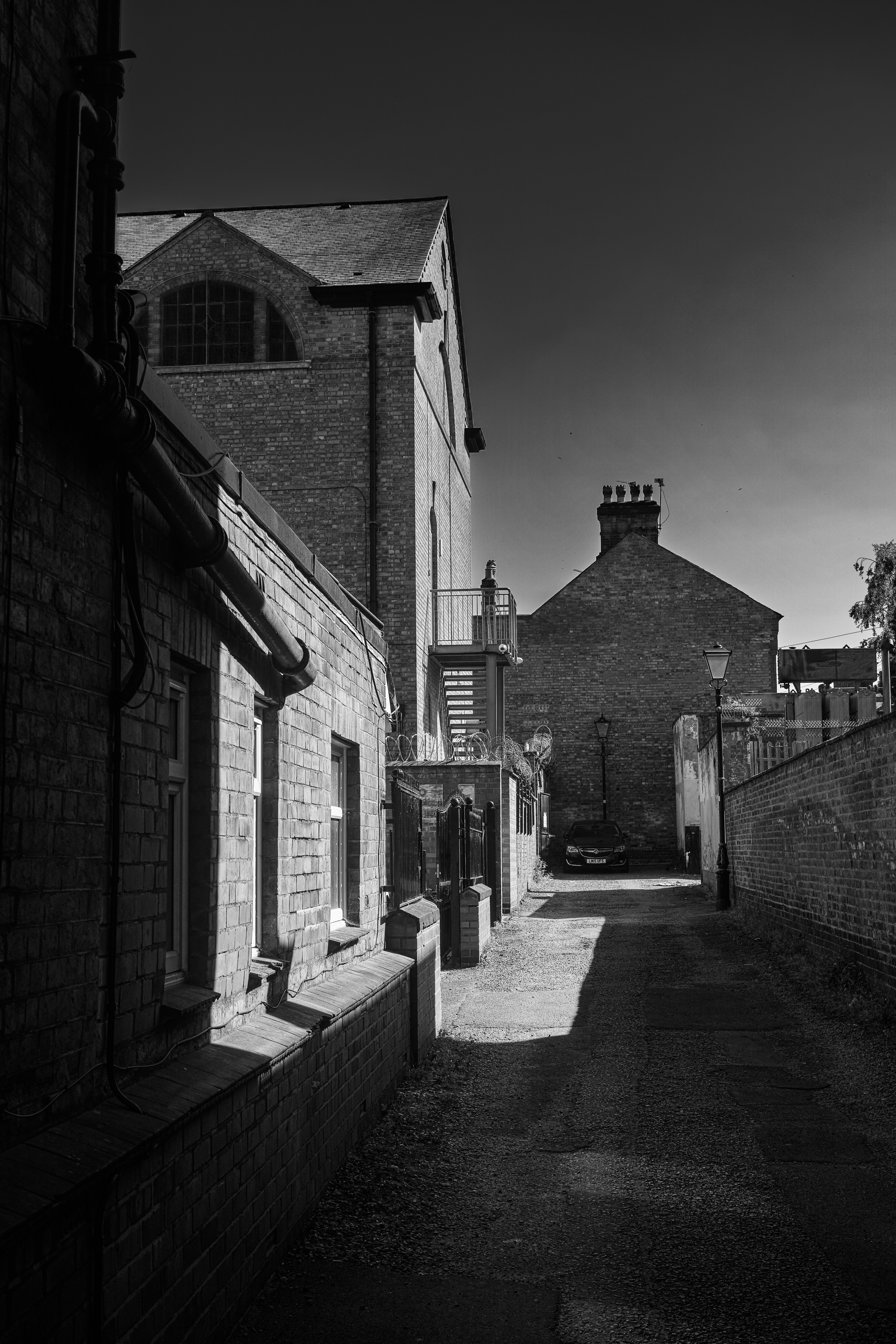 Monochrome alley scene narrowing toward a distant car. Sunlit walls cast stark shadows, emphasizing geometric brickwork and depth.