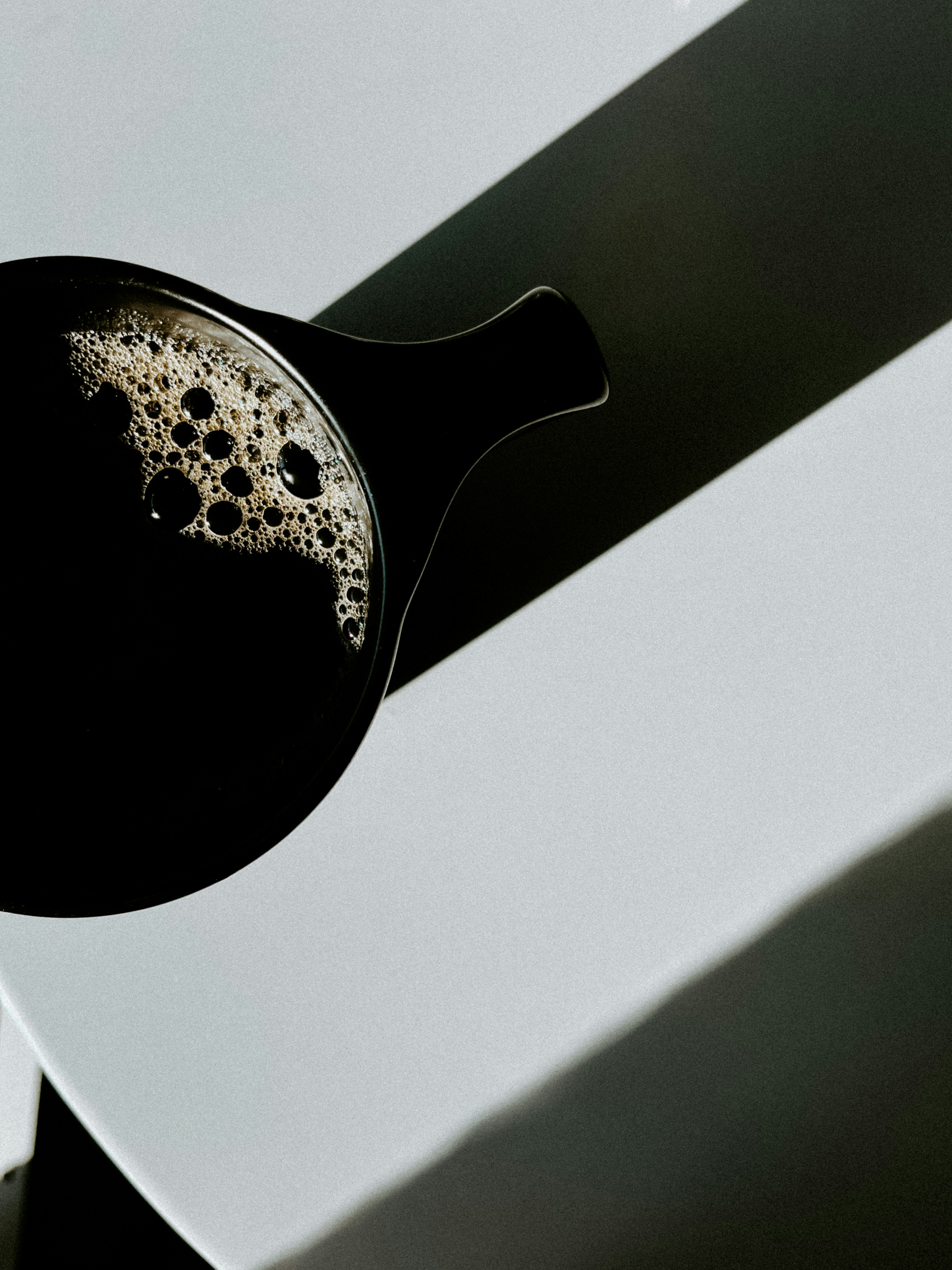 A minimalist photograph of a dark coffee vessel resting on a pale surface. Strong diagonal shadows create bold geometry across the frame.
