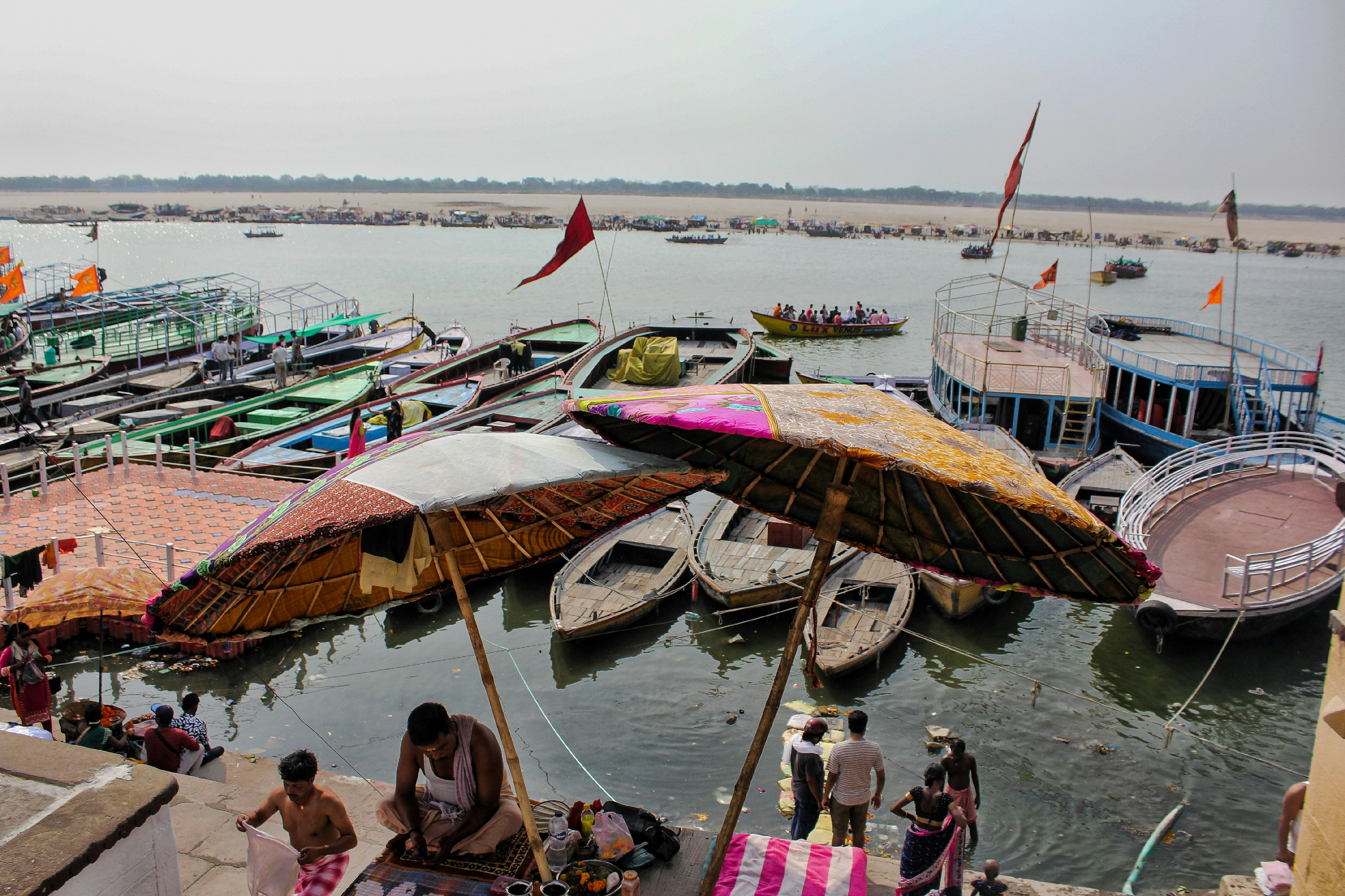 Boats docked along the Ganges River under large straw umbrellas, with people gathering on the steps.