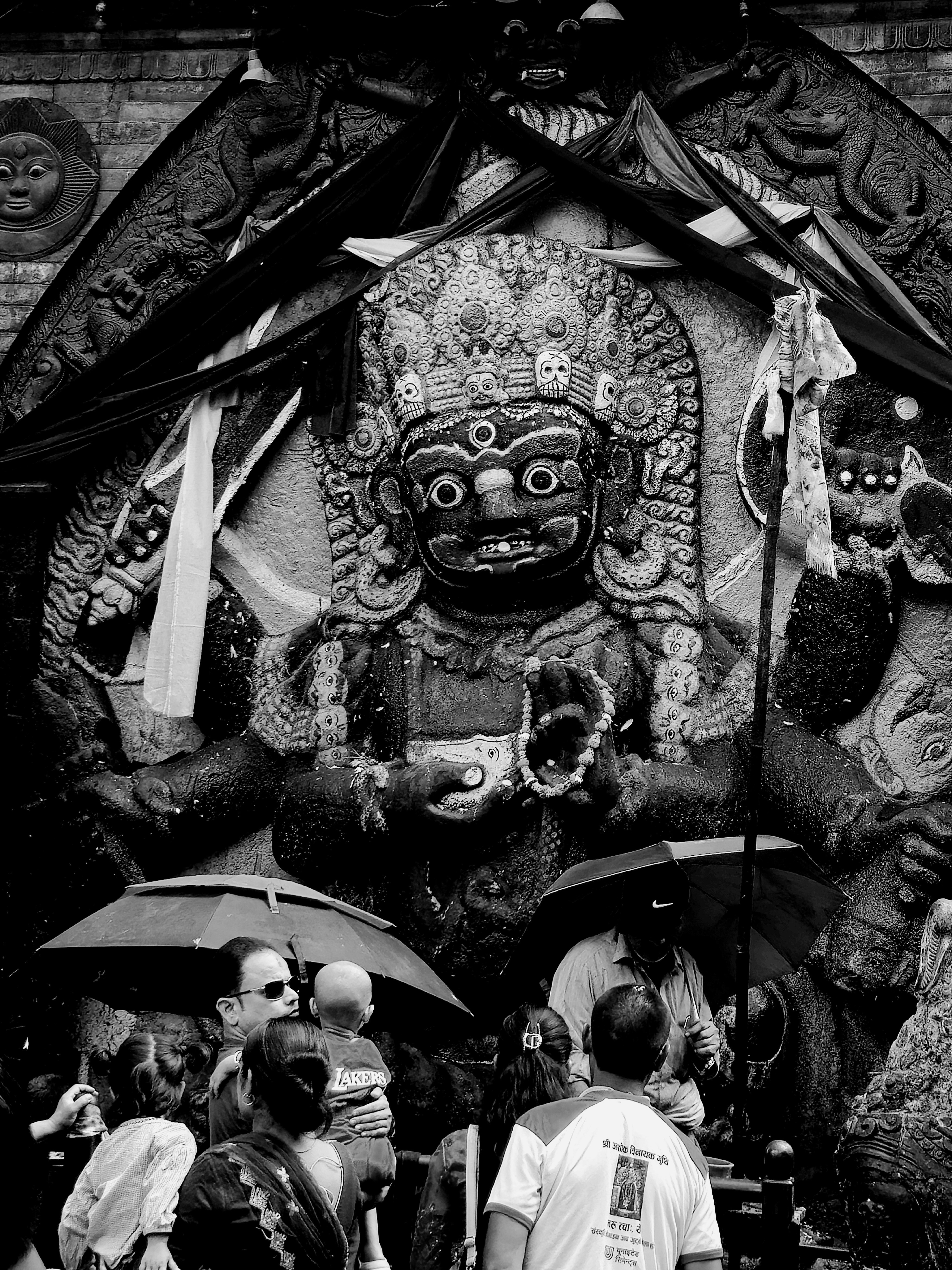 Intricate stone carving of a deity surrounded by onlookers, umbrellas, and cultural attire in a lively setting.