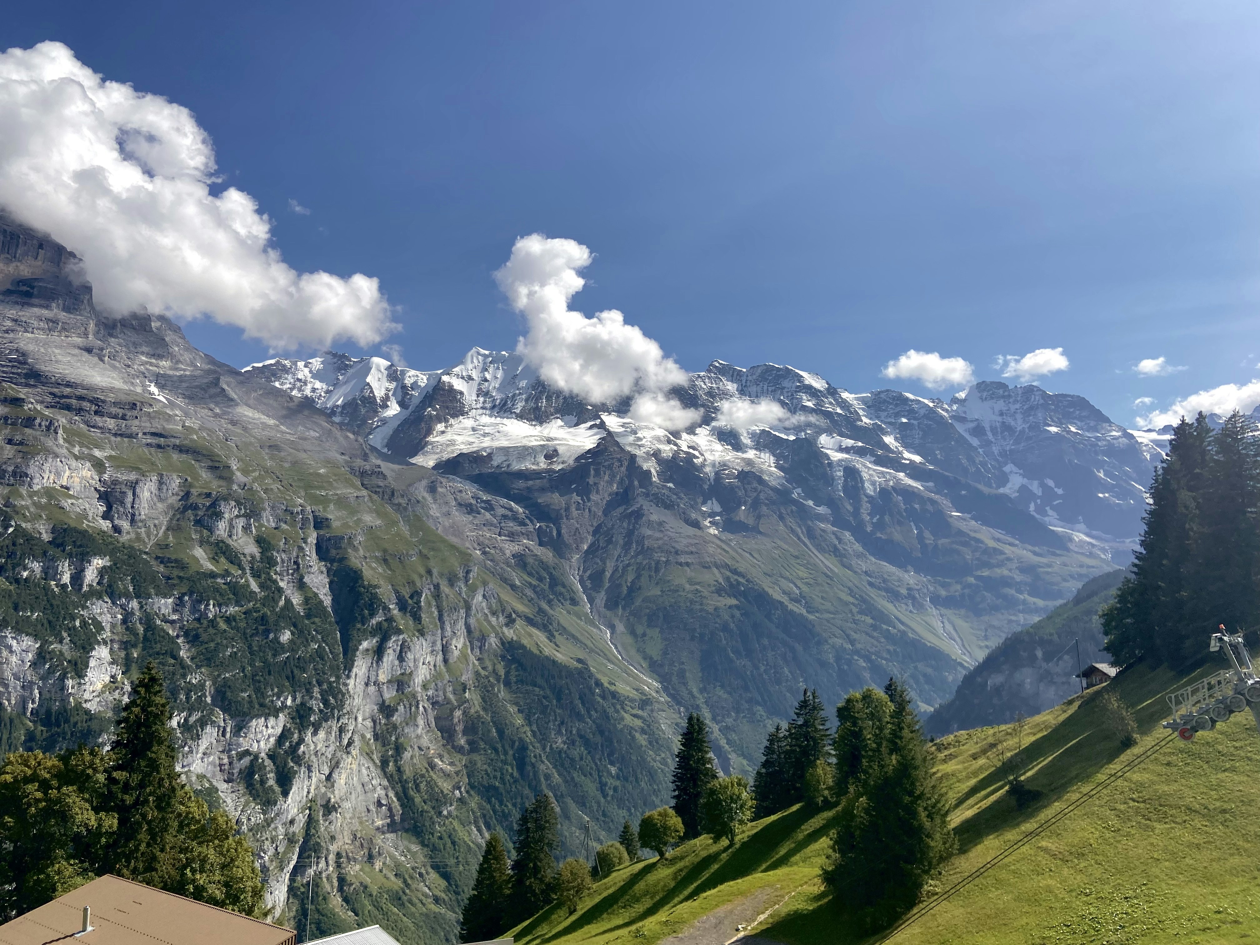 Vast mountain range under a clear blue sky, with clouds gently resting on the peaks and lush greenery in the foreground.