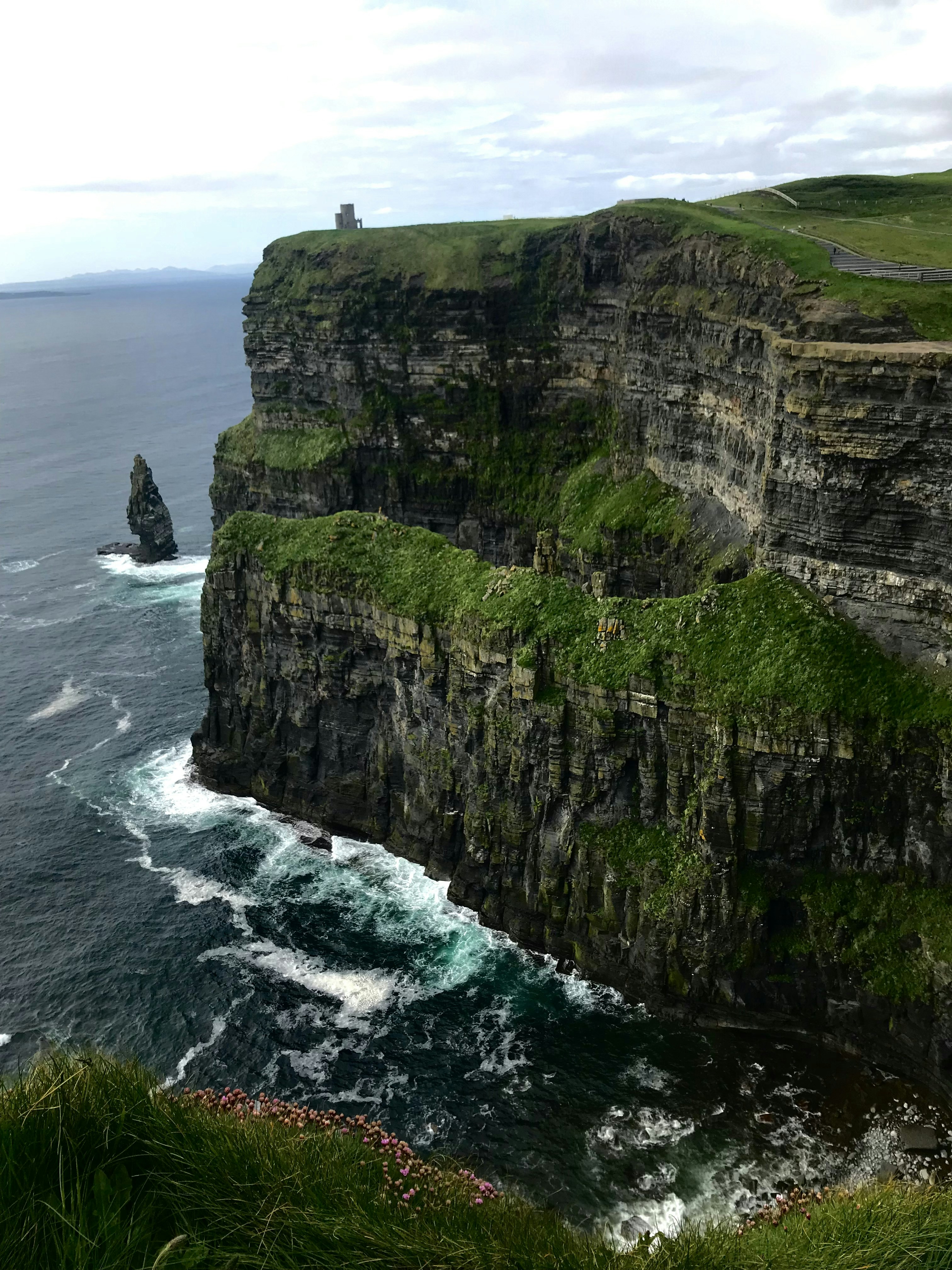 Majestic cliffs rise sharply from the Atlantic Ocean, adorned with lush greenery and dramatic rock formations. A distant tower stands sentinel over the rugged coastline.