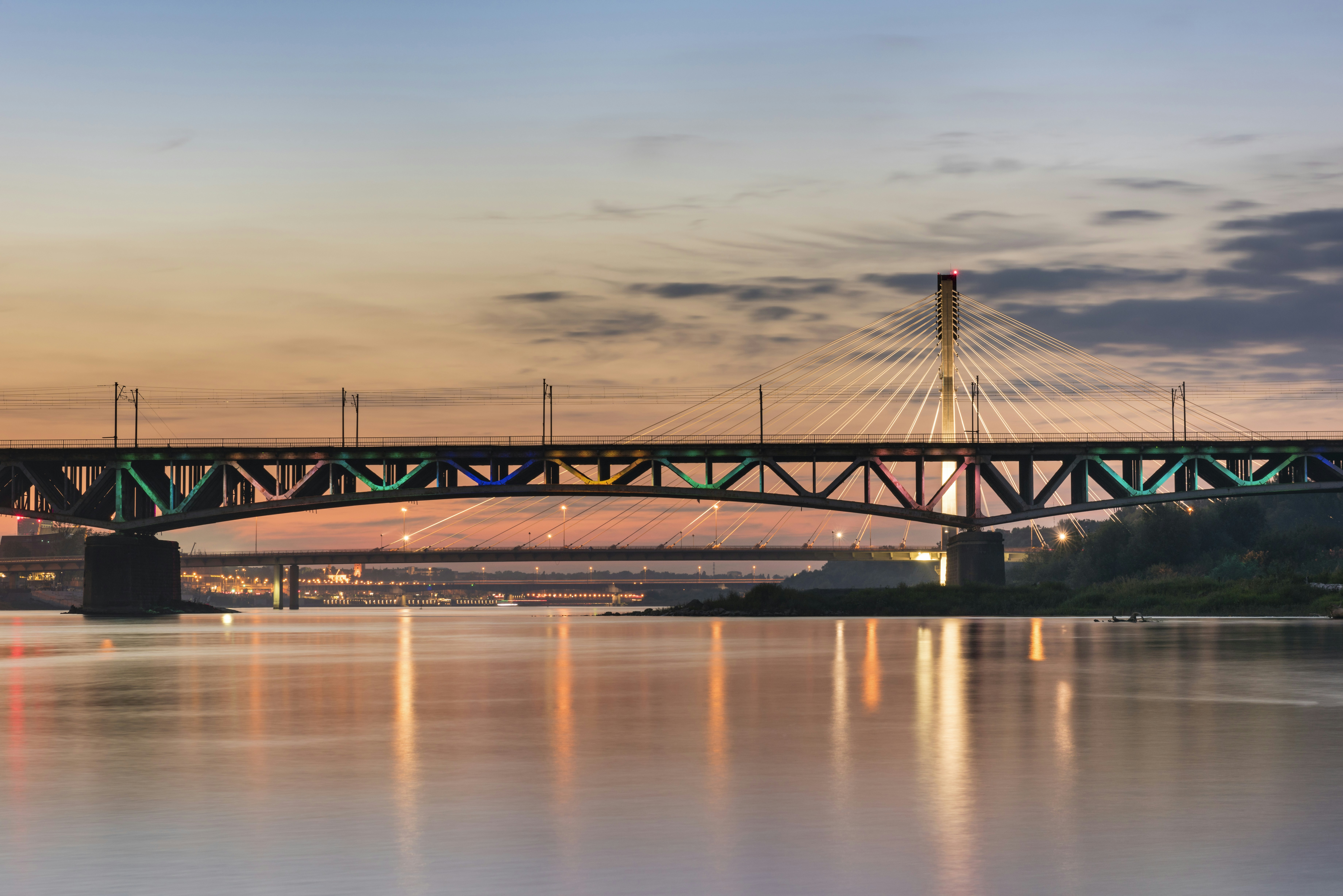Swietokrzyski Bridge spans the Vistula River at sunset, with soft pastel colors reflecting on the water.