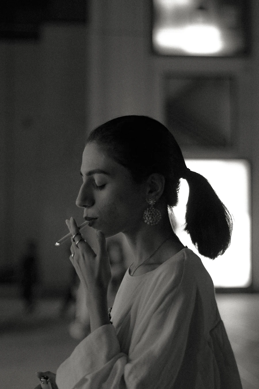 Black-and-white side profile of a woman with her eyes closed, holding a cigarette near her lips. Soft window light silhouettes her ponytail and earrings in a dim interior.