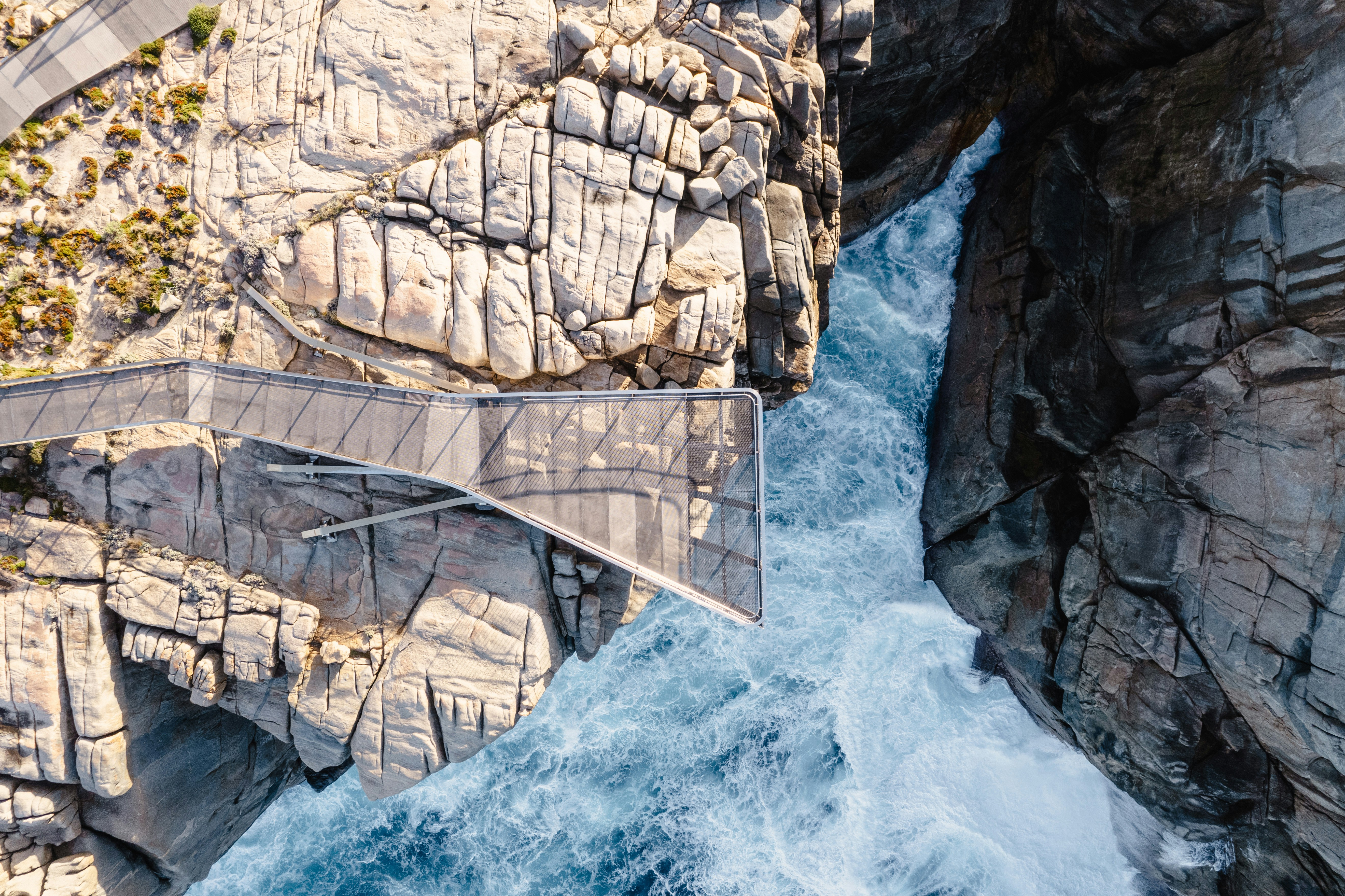 Aerial view of a geometric walkway spanning rugged cliffs above churning blue waters.