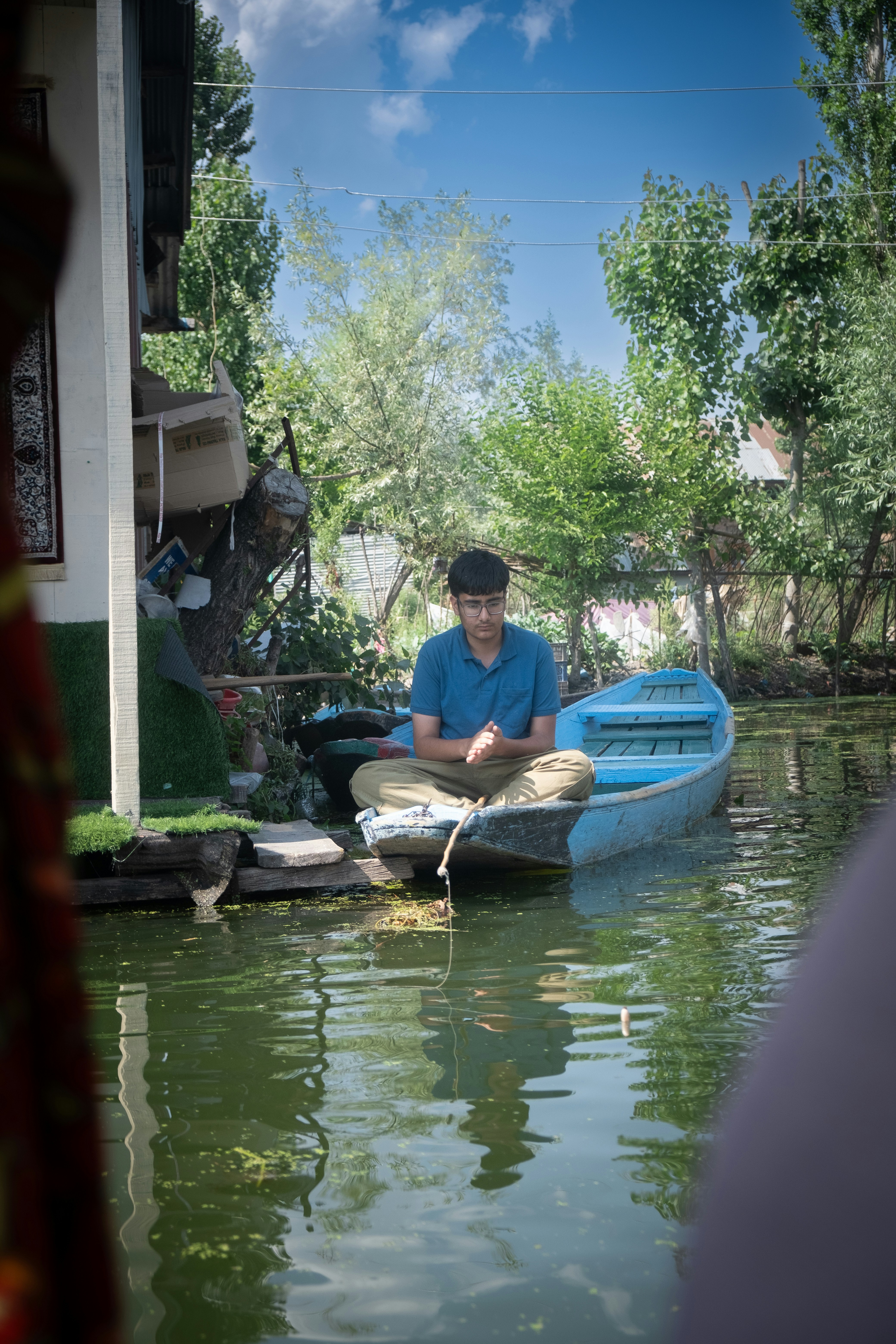 Blue Boat Solitude: A man meditates on a sunlit canal beside a riverside home.