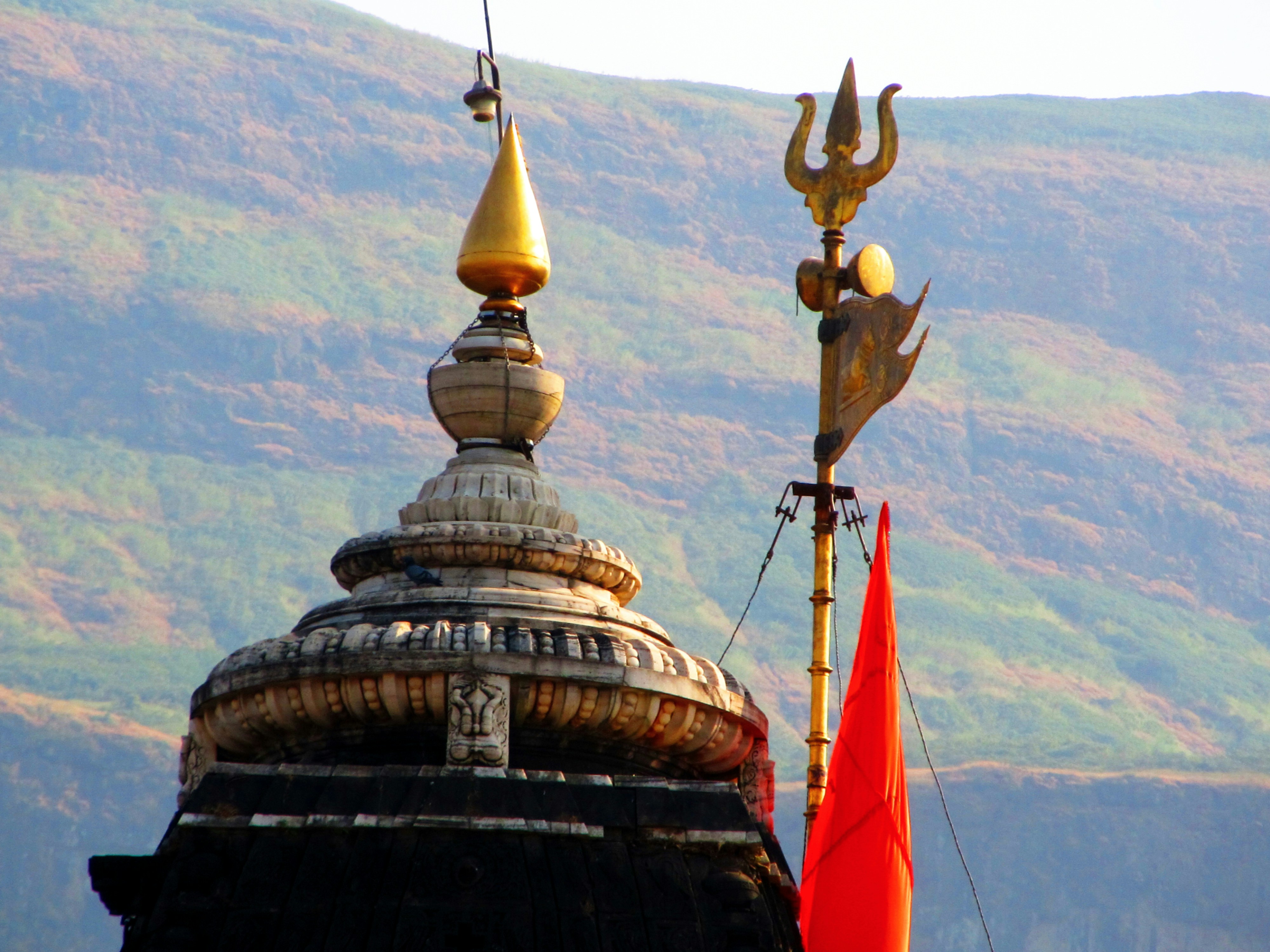 Intricate temple spire adorned with a golden finial and flag, set against a backdrop of rolling hills. The craftsmanship reflects cultural heritage.