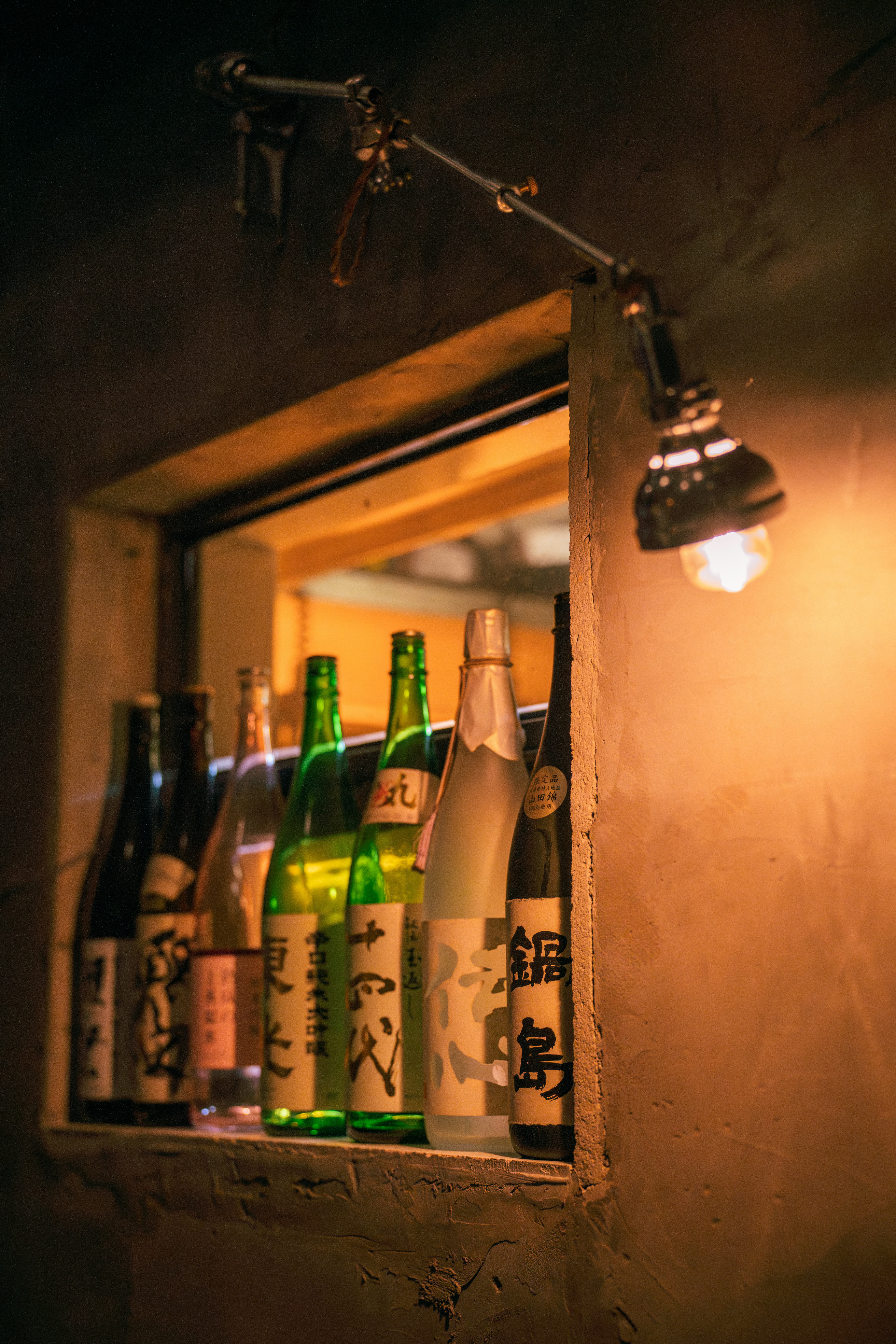 A collection of sake bottles displayed in a window, softly illuminated by a hanging light, showcasing their unique labels and colors against a textured wall.