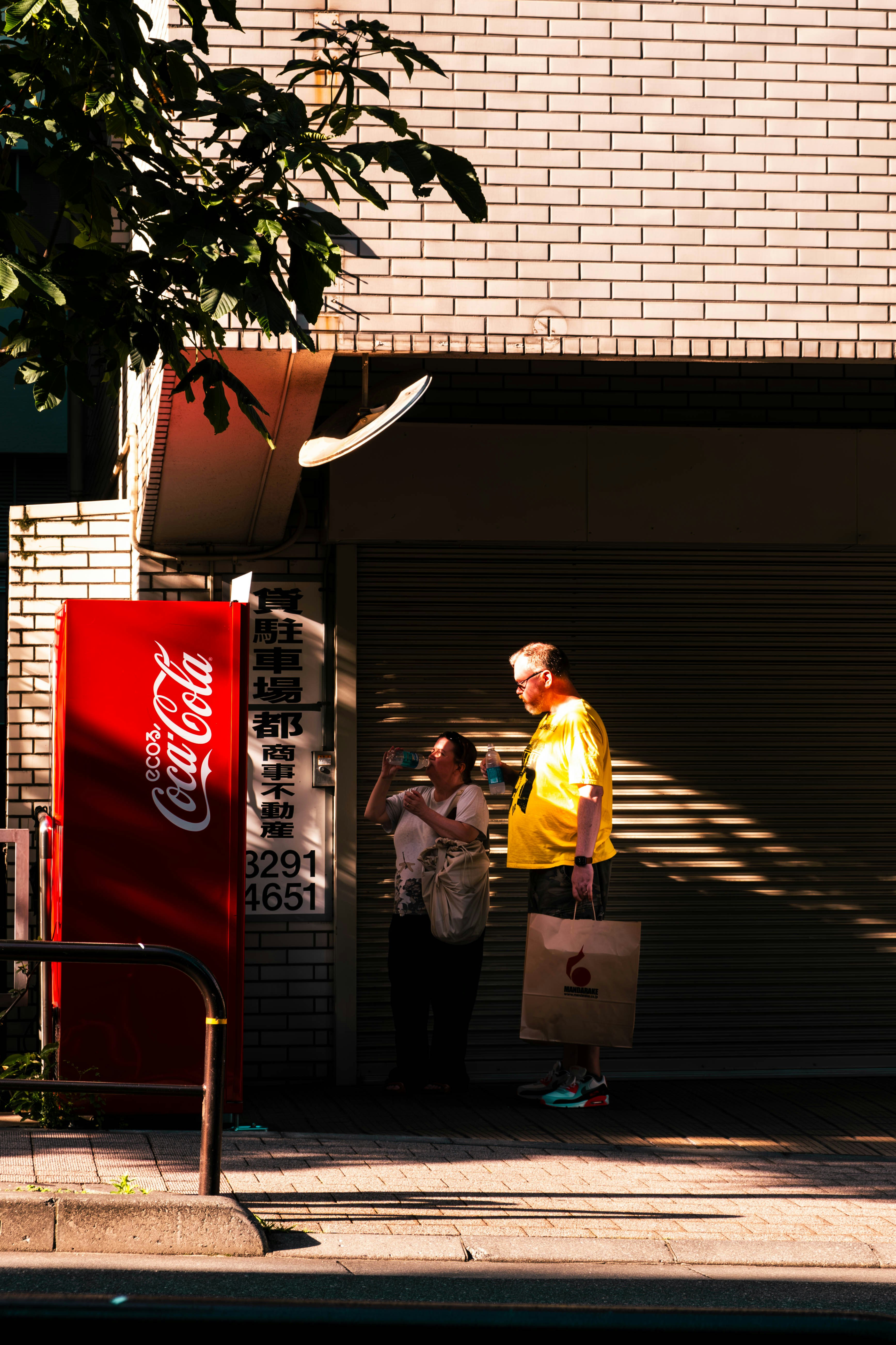 Sunlit Exchange by the Brick Wall: A Pedestrian Snapshot Near a Coca-Cola Vending