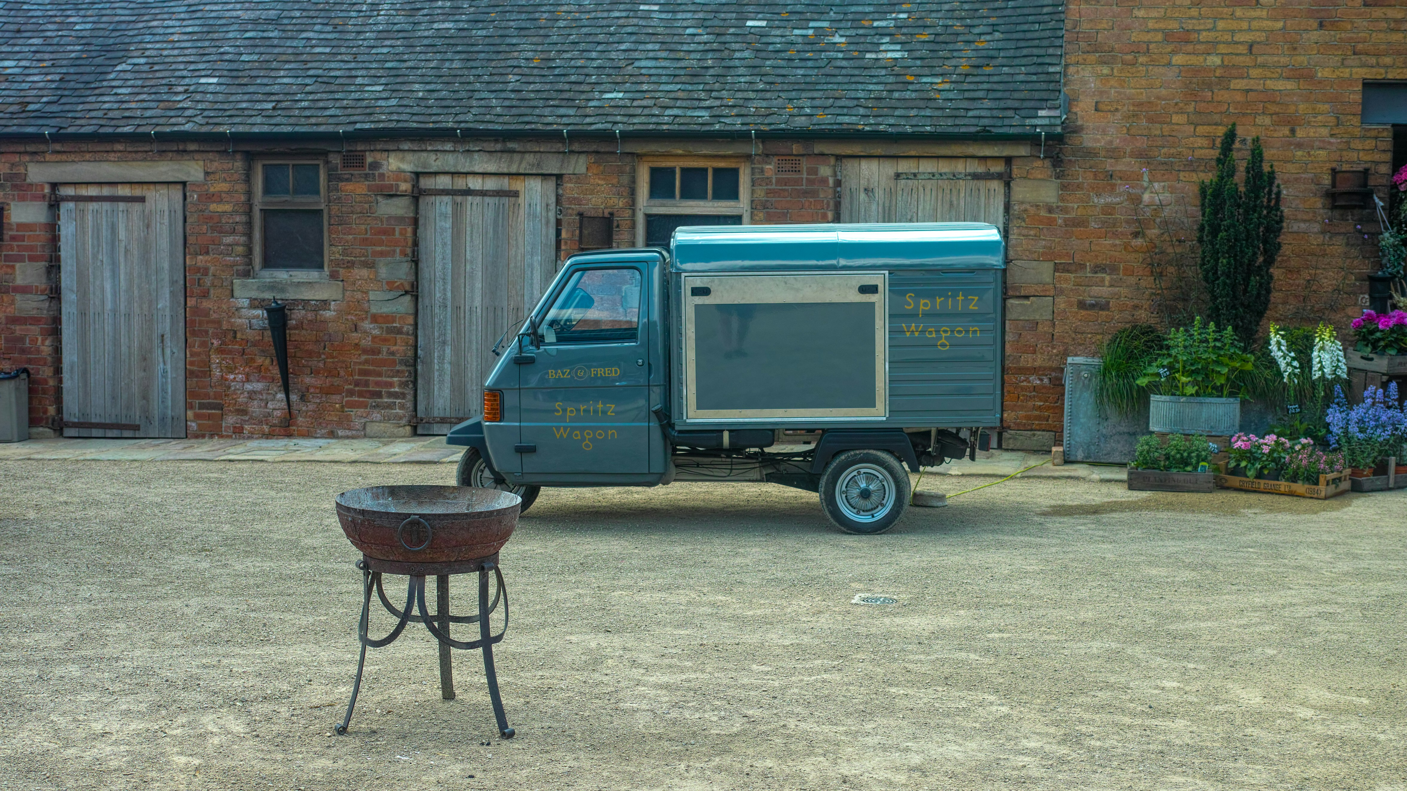 A mint-blue micro-camper is parked in a gravel yard beside a brick building. A metal fire pit sits in the foreground, adding domestic contrast.