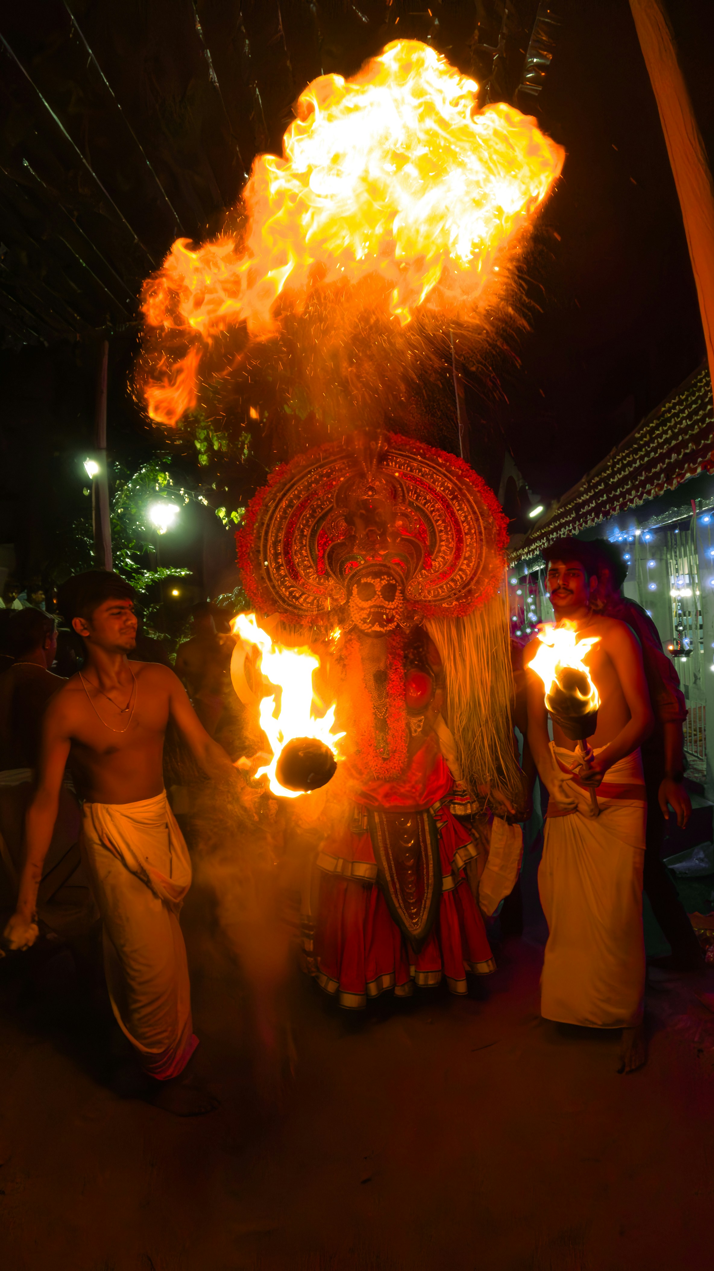 The image captures a captivating fire-breathing performance. The central figure, dressed in an elaborate costume with a large, ornate headdress, is breathing a large plume of fire into the air. Two other individuals are assisting with the performance, one holding a flaming object. The scene is set at night, illuminated by artificial lighting, suggesting it might be part of a cultural or religious festival. | A group of people standing around a fire pit