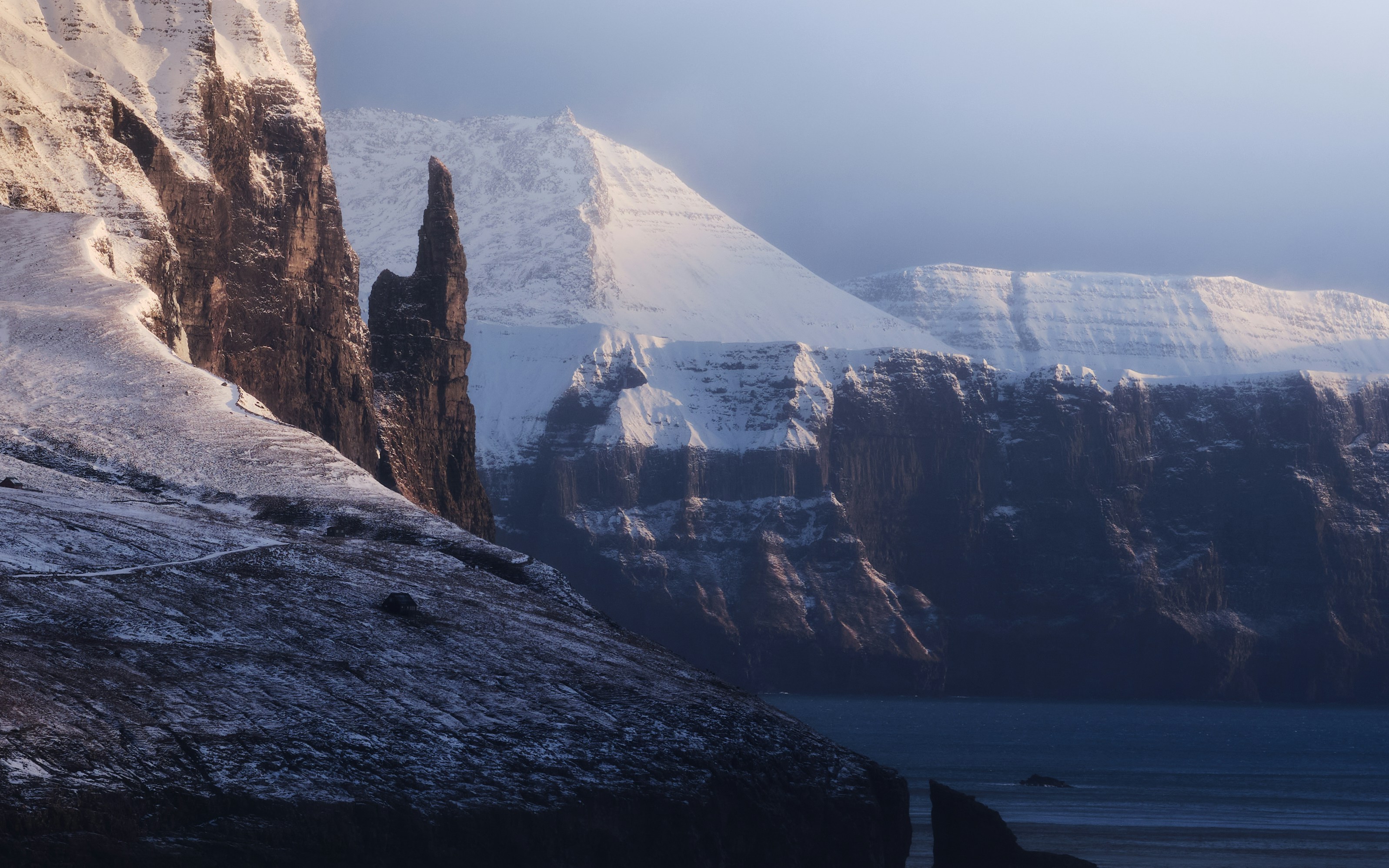 Sunlit snowy mountain with a sharp rock formation rising beside a serene body of water.