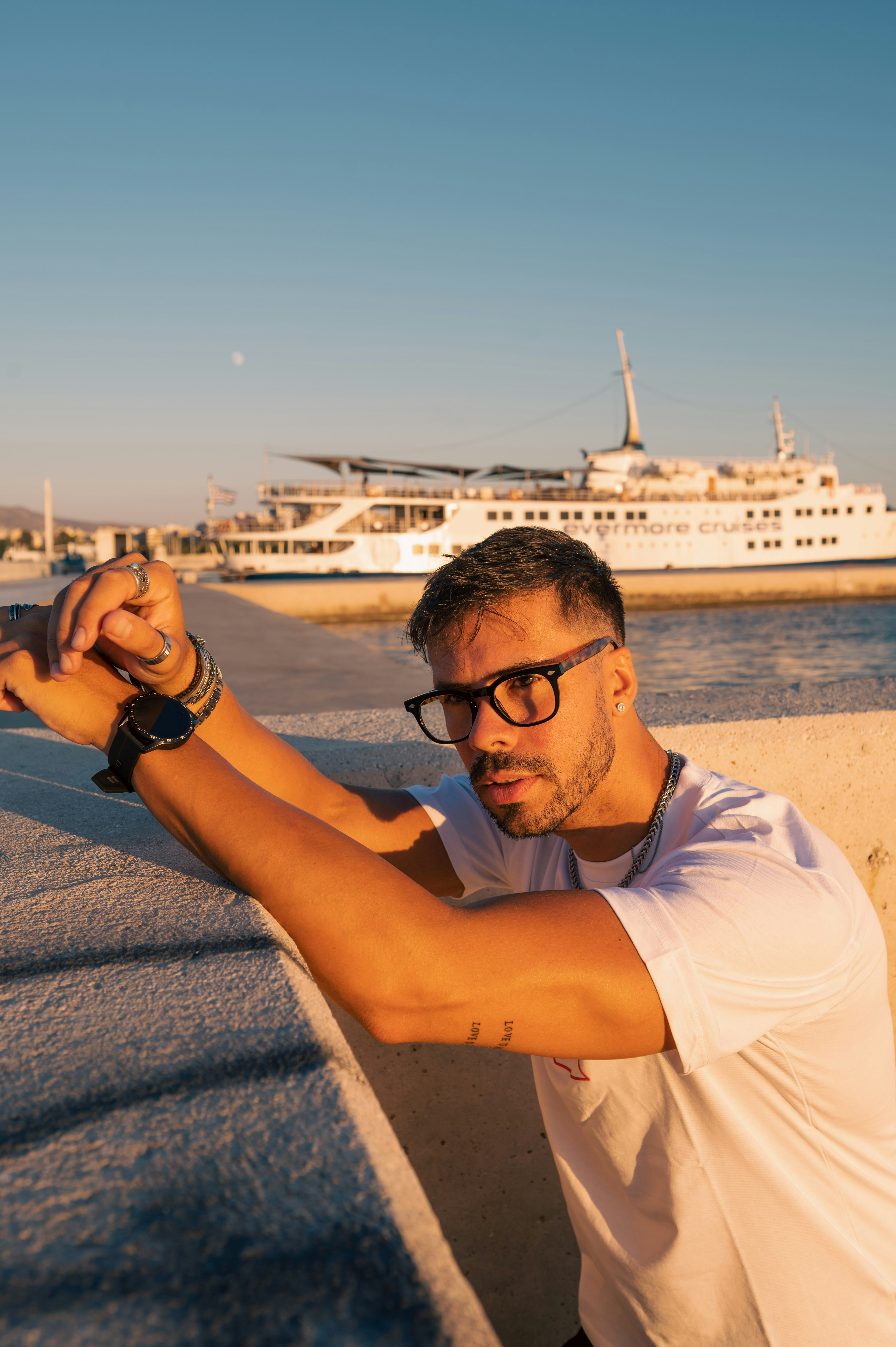 A stylish man relaxes by the waterfront, sporting sleek black-framed glasses that accentuate his sharp features. His casual yet chic outfit, complemented by his accessories, matches the serene coastal backdrop. The glasses add a touch of intellectual charm, enhancing his overall modern and laid-back look.