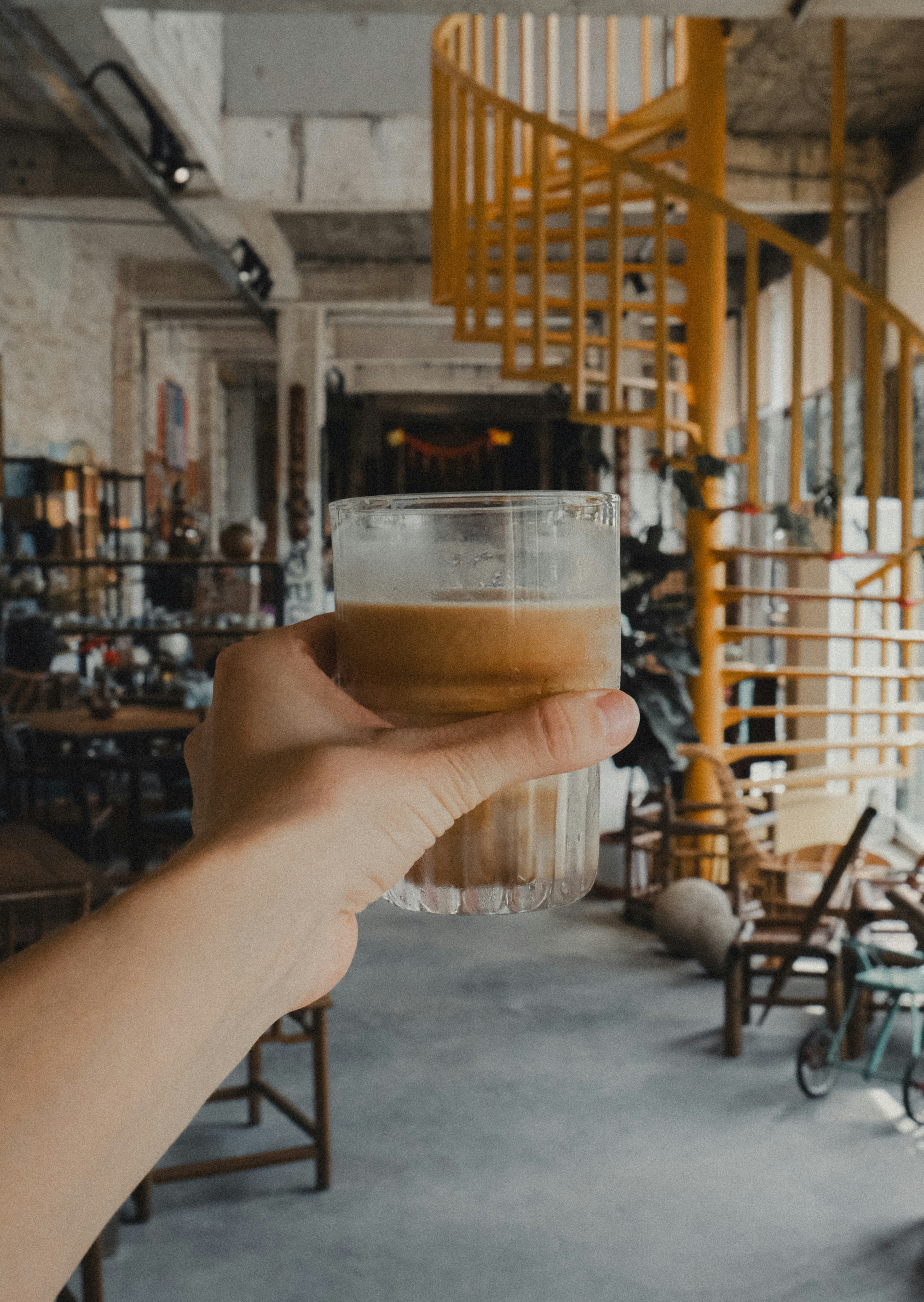 A hand holds a frothy iced coffee in a textured glass in a spacious industrial café. The scene features a bright yellow spiral staircase and softly blurred patrons.